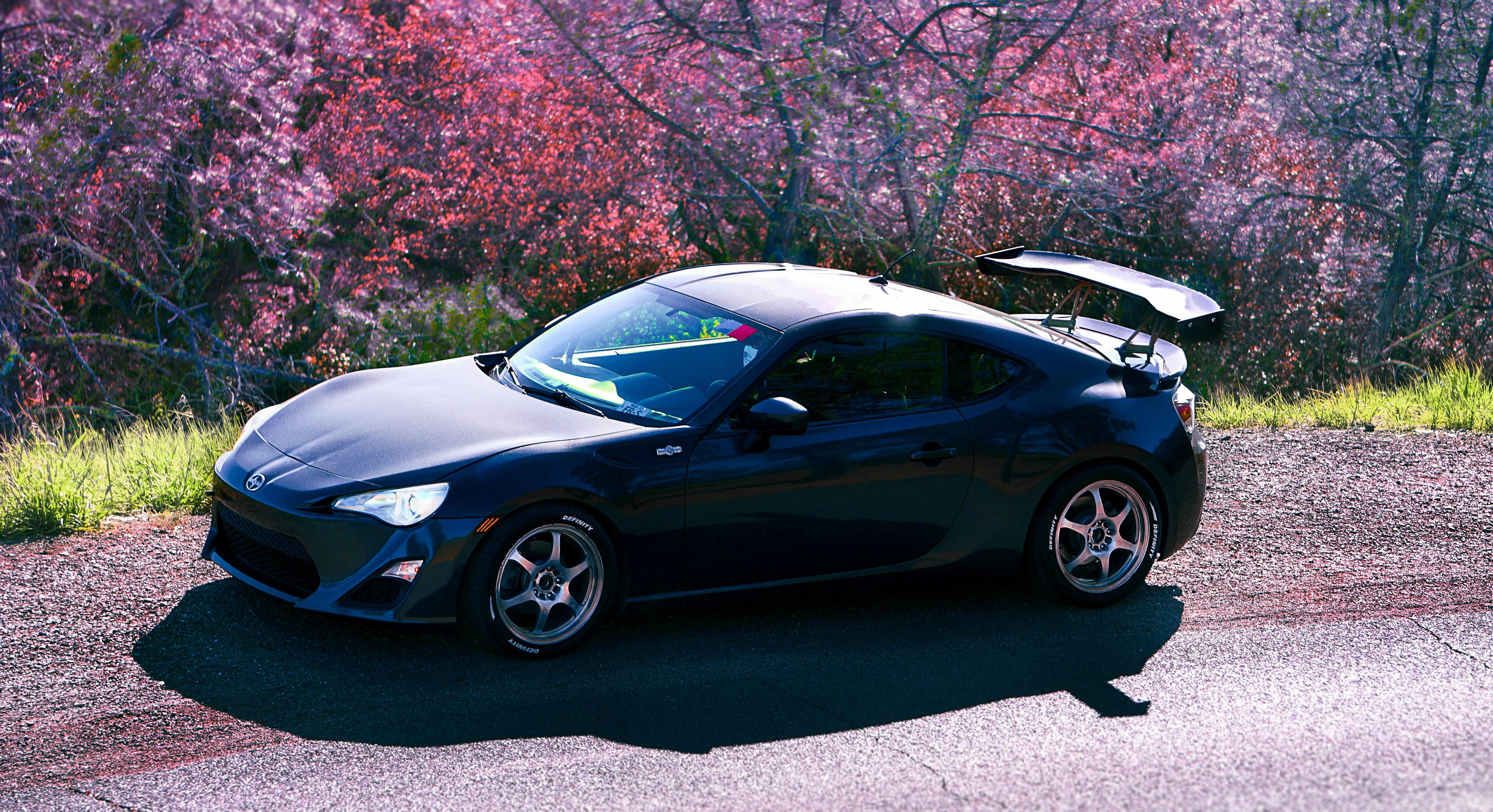 A sleek black sports car parked on a winding road surrounded by vibrant pink blossoms, showcasing its aerodynamic design and sporty features.