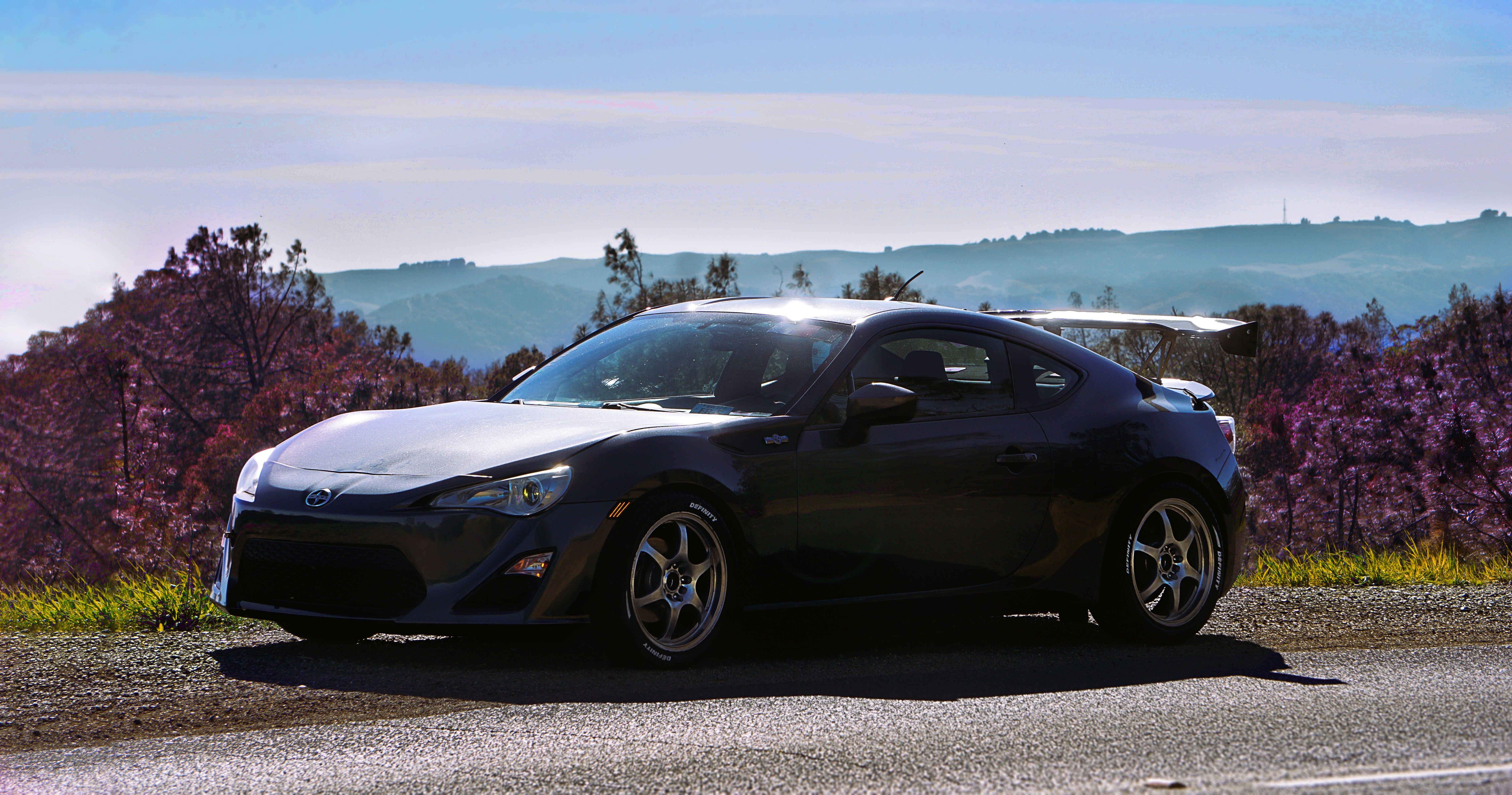 Sleek black sports car parked on a winding road, surrounded by vibrant purple foliage and distant hills under a clear sky.