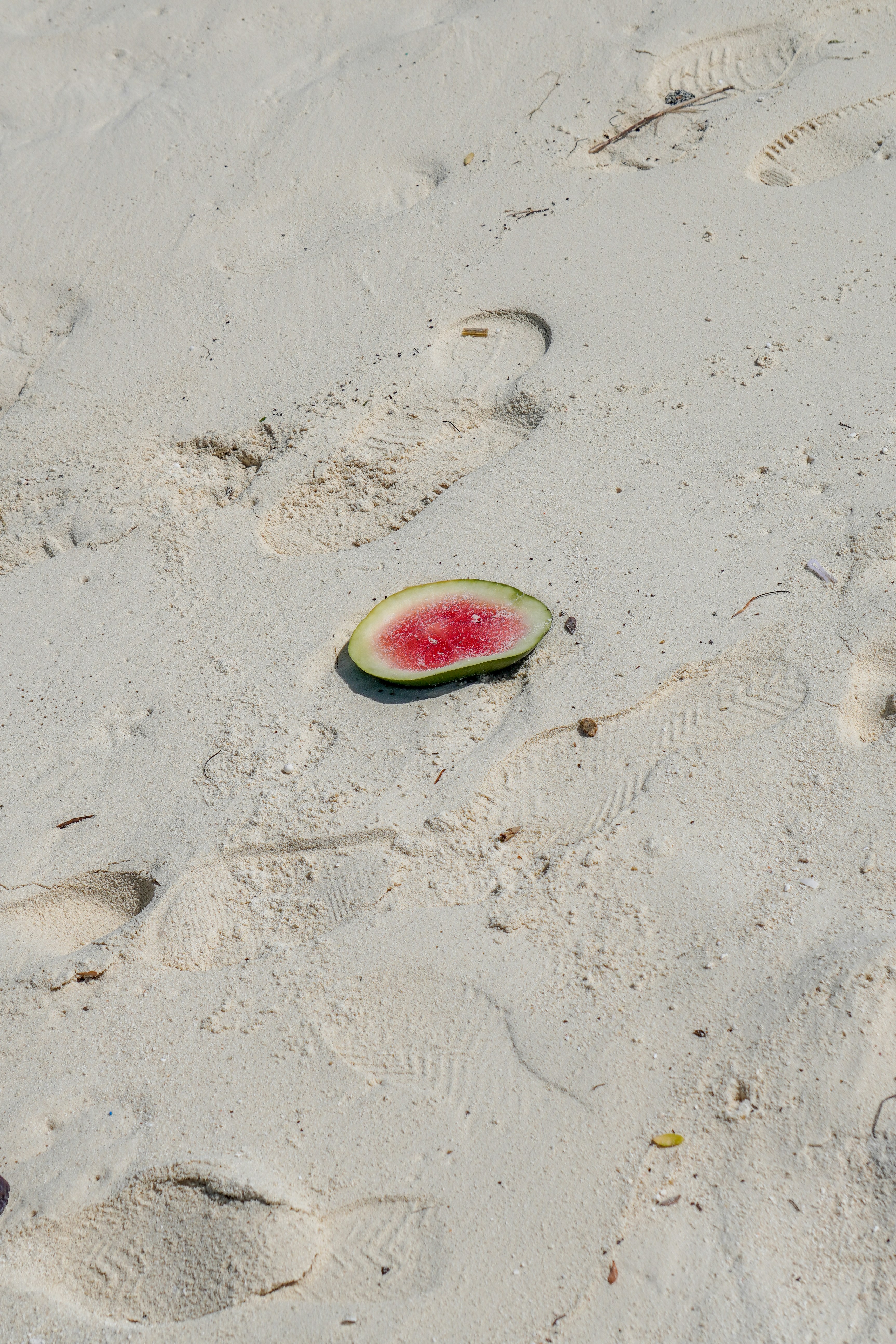 Watermelon slice resting on sunlit beach sand, surrounded by footprints. The scene captures a moment of casual summer enjoyment.