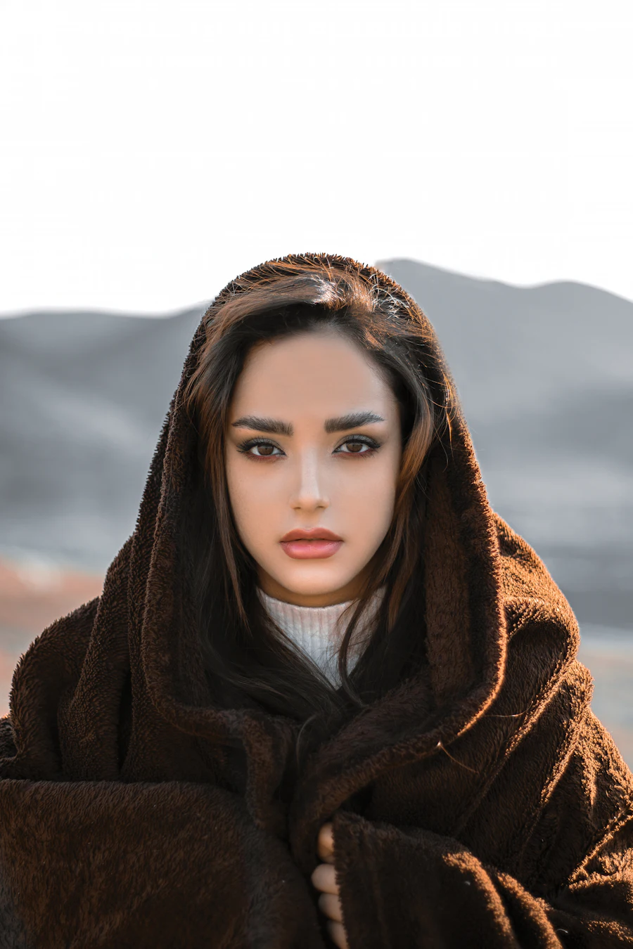Centered portrait of a woman wearing a dark brown hooded wrap, with a soft blurred mountain backdrop and bright sky behind her. Her face and eyes are the clear focal point.
