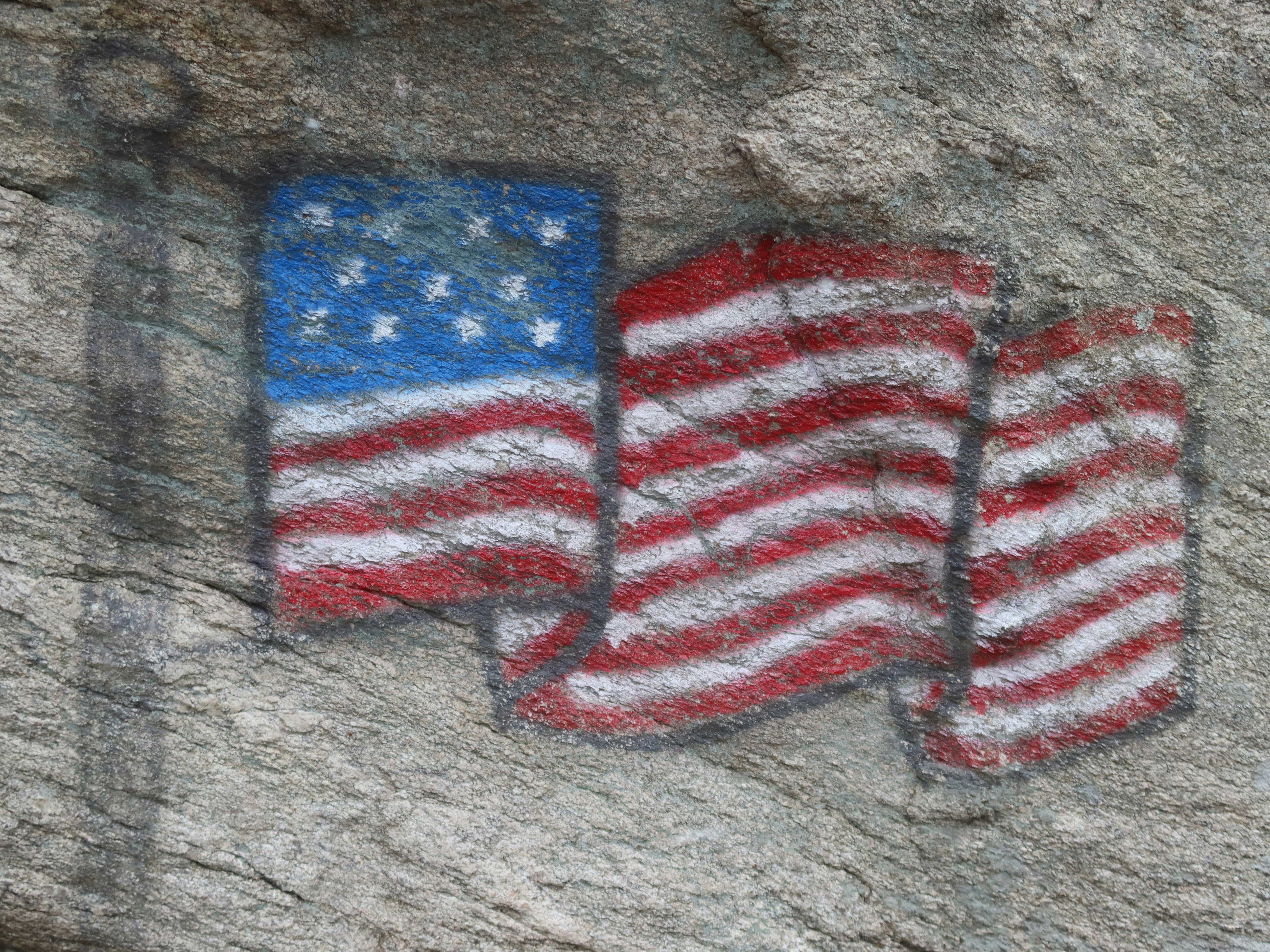 A weathered rock surface features a painted American flag, showcasing vibrant colors despite the rough texture. The artwork symbolizes resilience and national pride.