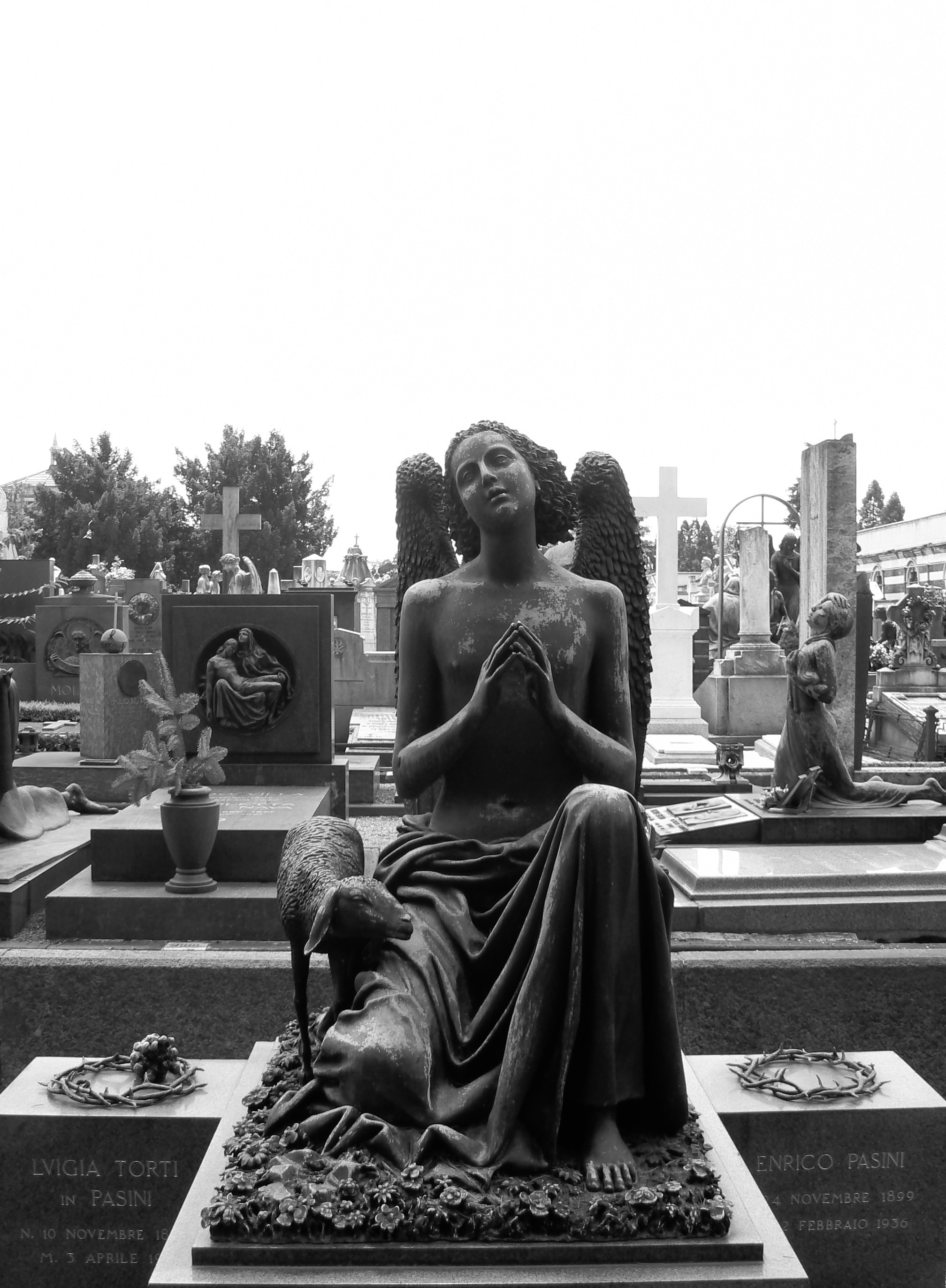 Monochrome statue of a seated angel with hands clasped, surrounded by weathered tombstones in a quiet cemetery.