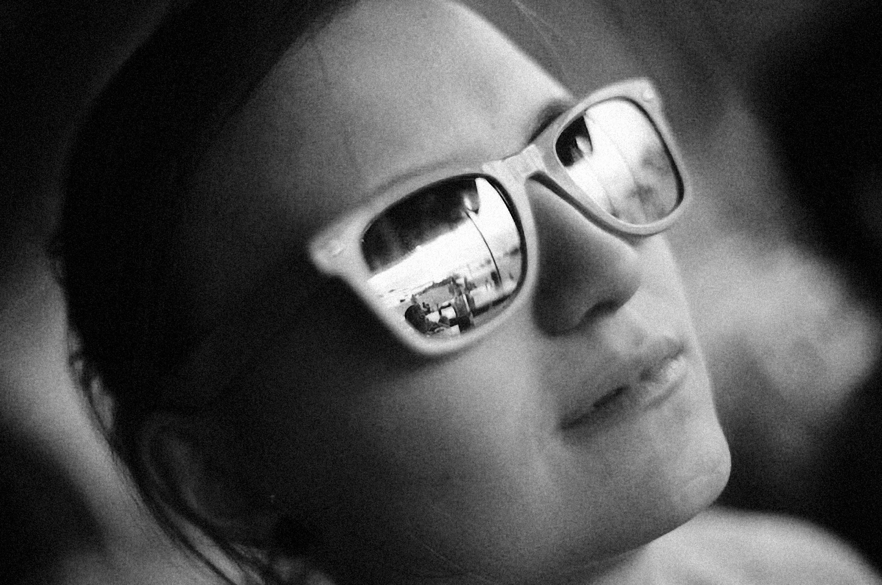 A woman gazes thoughtfully while wearing reflective sunglasses that capture a scene behind her. The monochrome tones add a nostalgic touch.