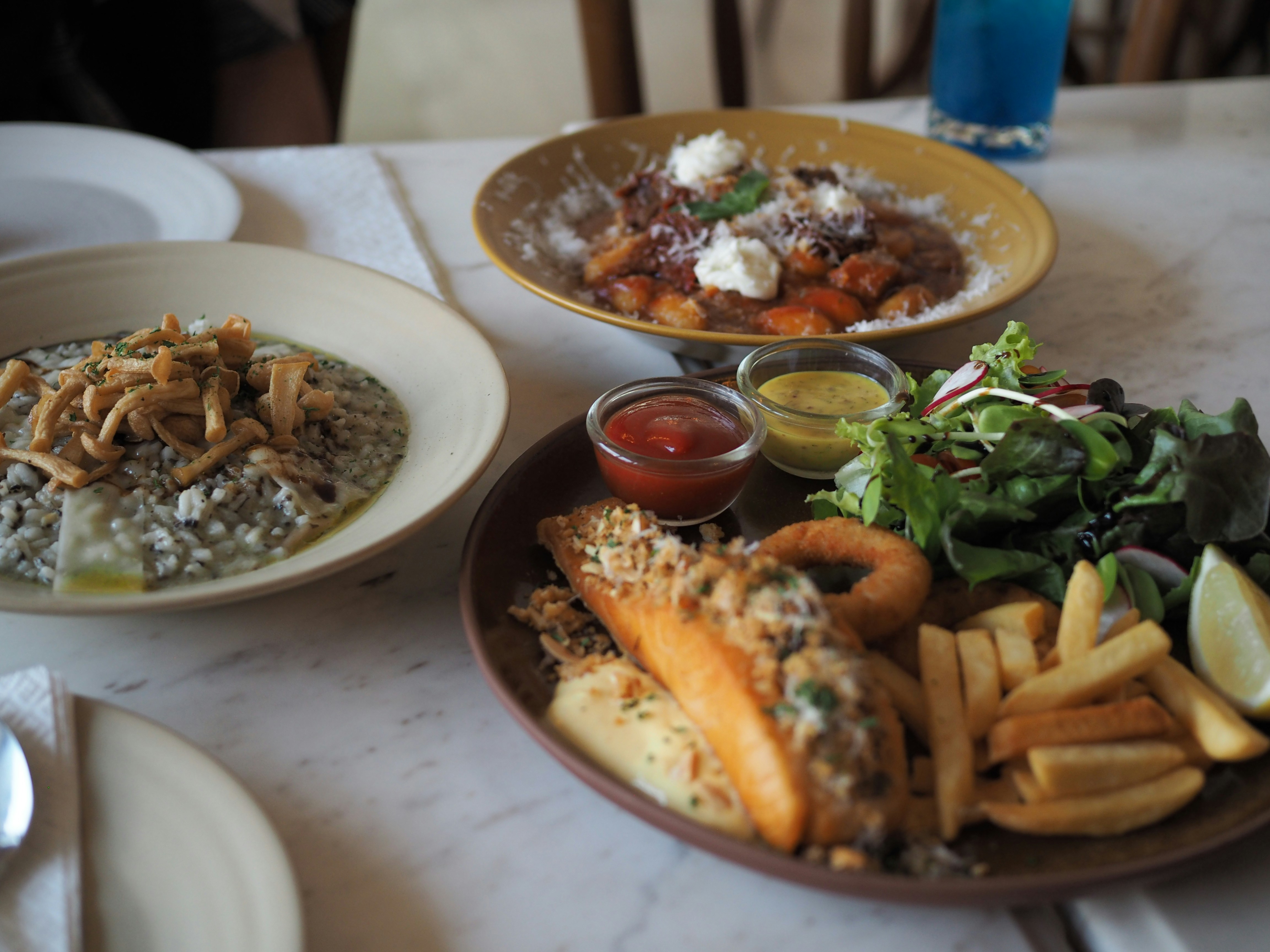A table of assorted dishes with a pasta plate, a brown plate of fried items and greens, and a small bowl of sauces in the center; a blue ice-in-glass in the background. A casual dining spread ready to share.