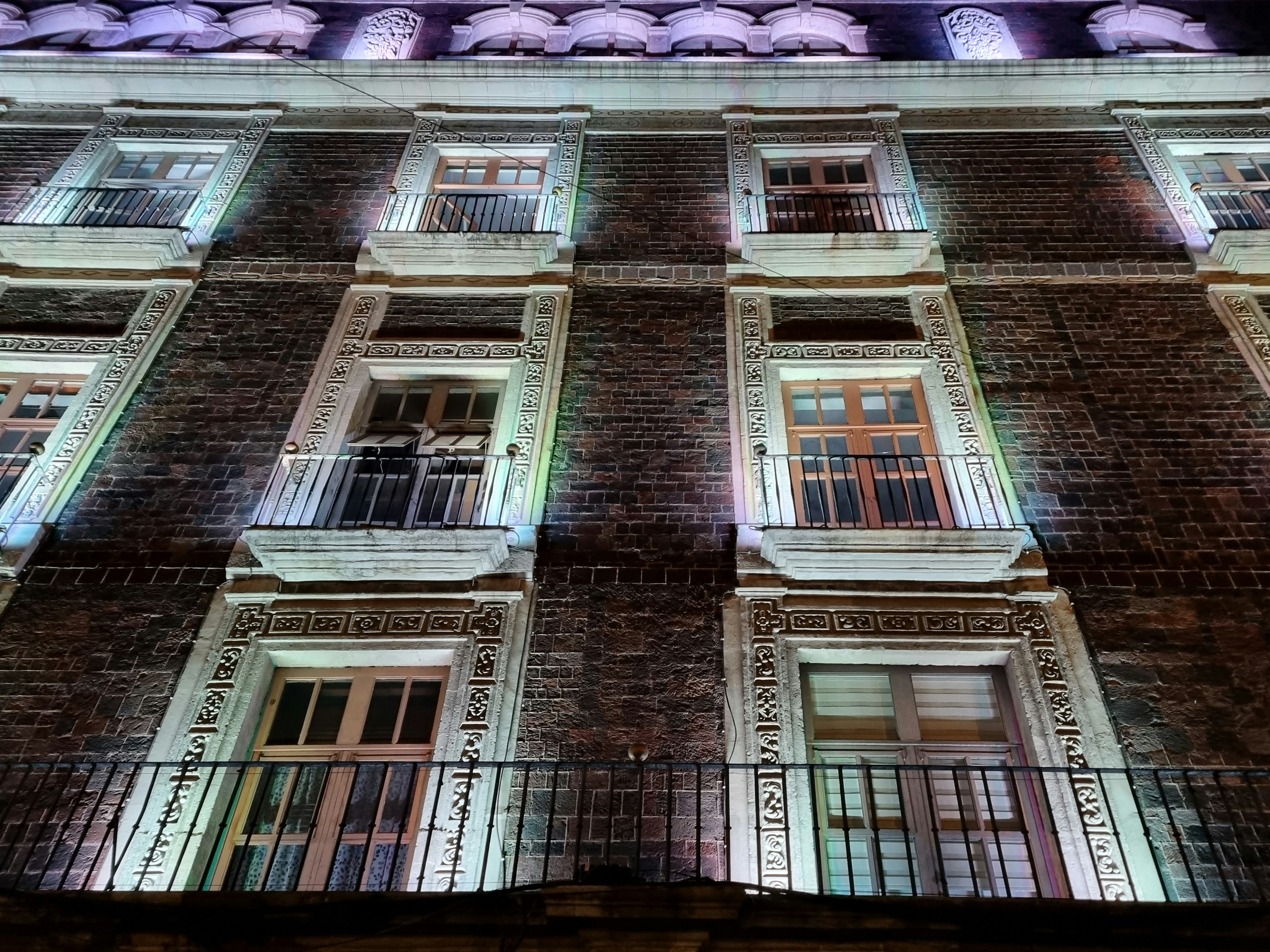 Night-lit brick facade with ornate window frames and wrought-iron balconies bathed in purple and green neon accents.