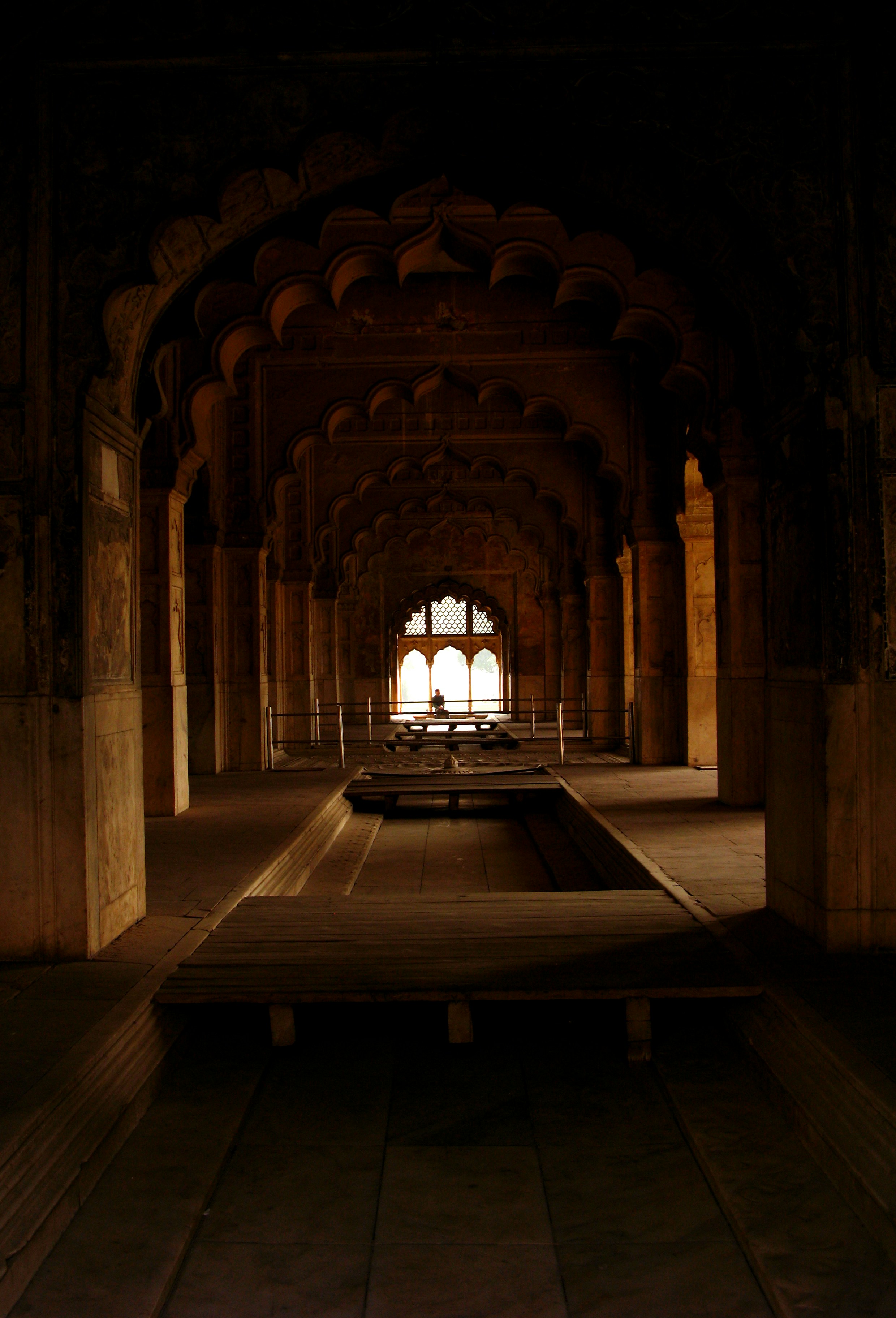 Dim interior corridor of carved arches leads to a sunlit doorway, with warm amber tones accenting the stone columns.