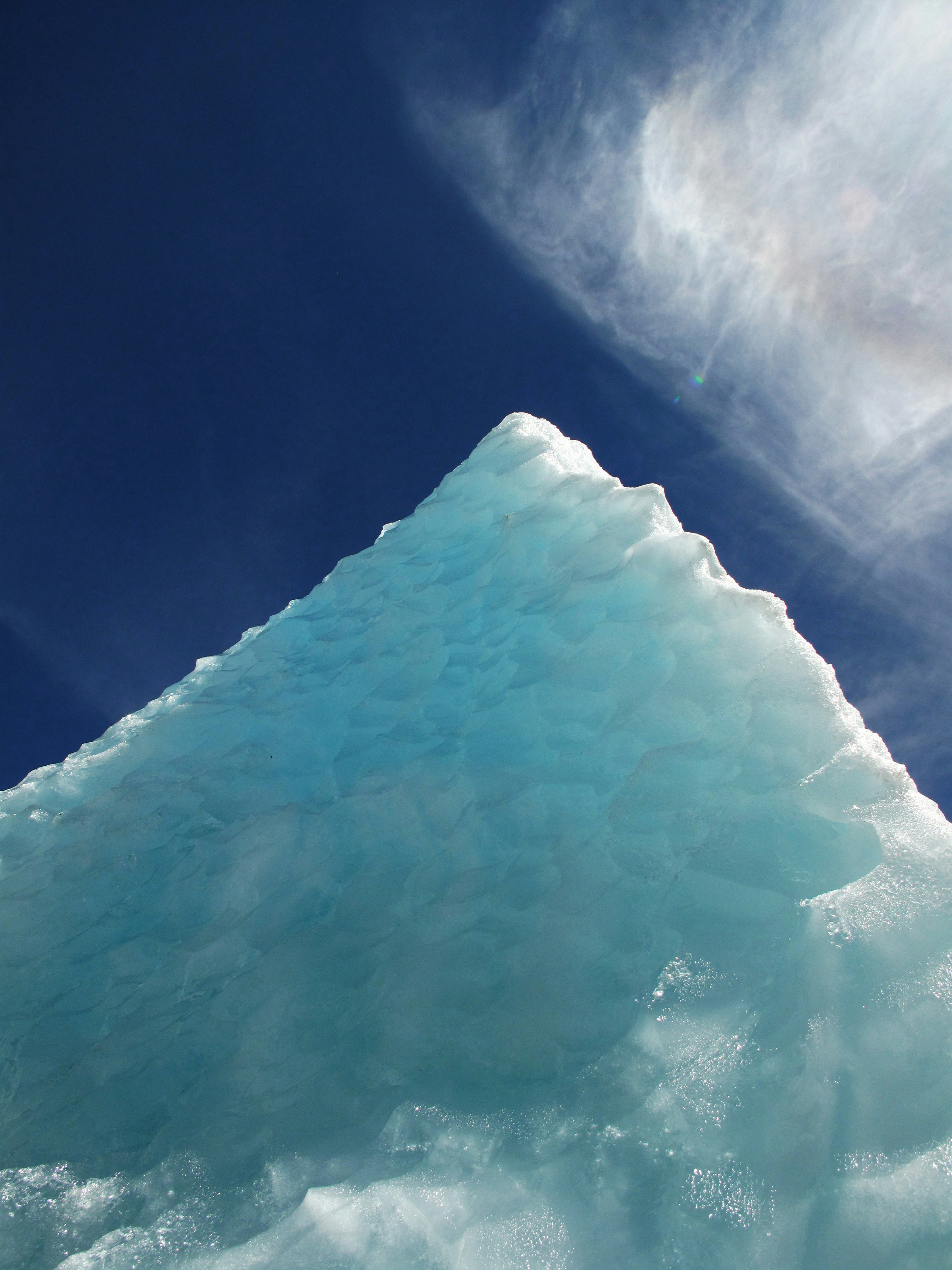 A towering ice formation reaching towards the sky, illuminated by sunlight, with a clear blue backdrop and wispy clouds above.