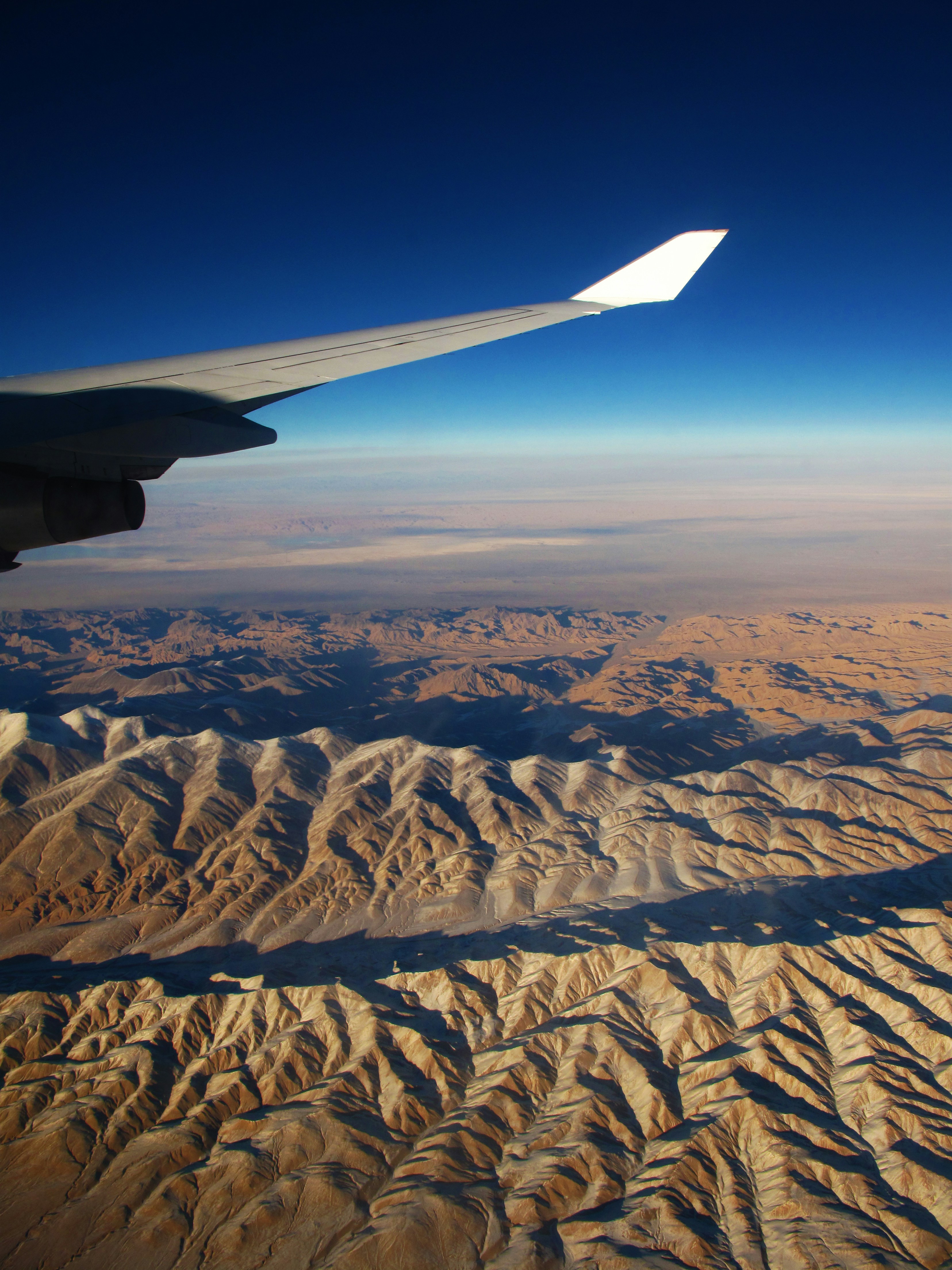 Airplane wing gliding above vast, undulating desert mountains under a clear blue sky.