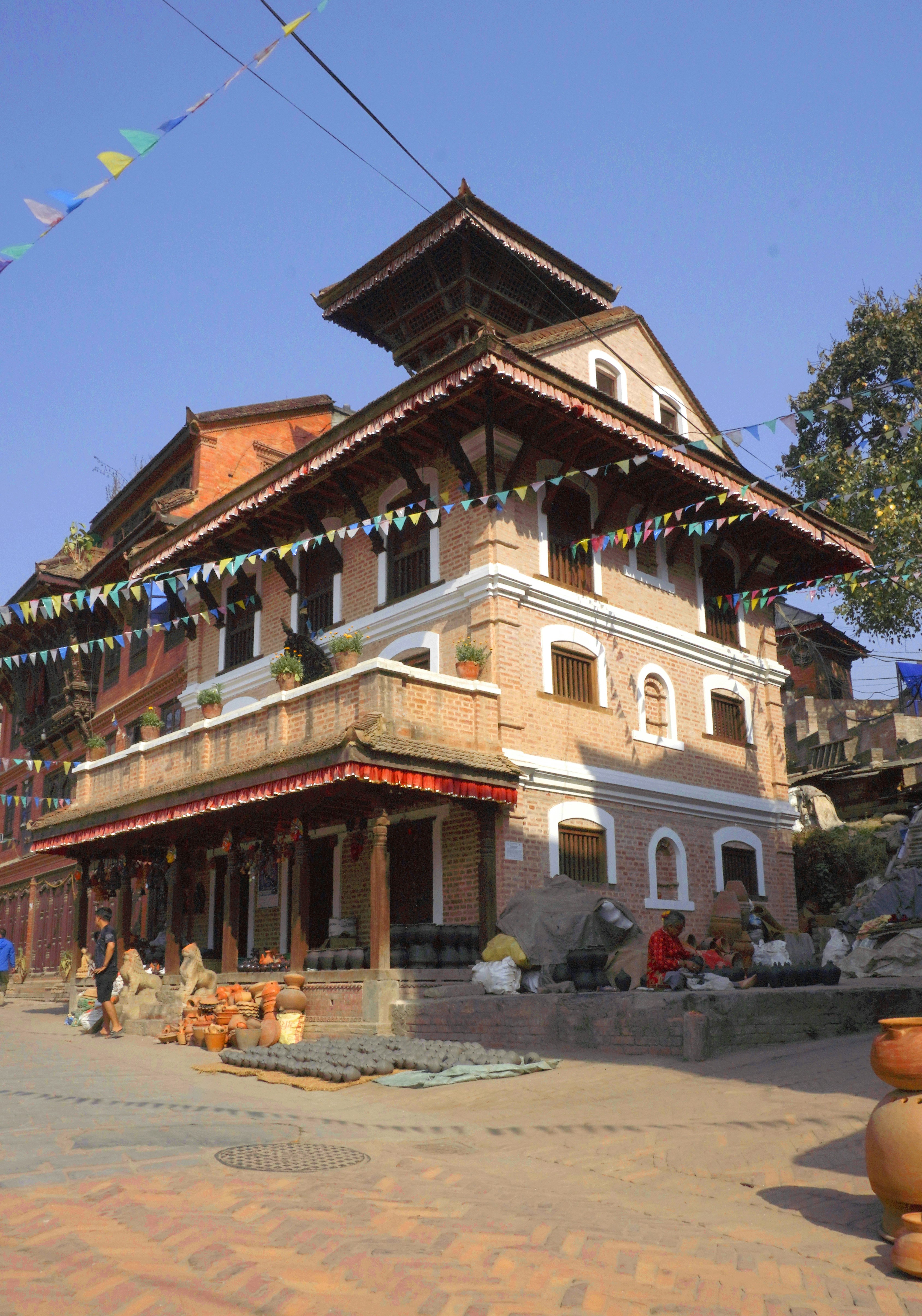 Historic multi-story building adorned with colorful prayer flags, showcasing intricate brickwork and traditional Nepali design. Visitors engage in local crafts nearby.