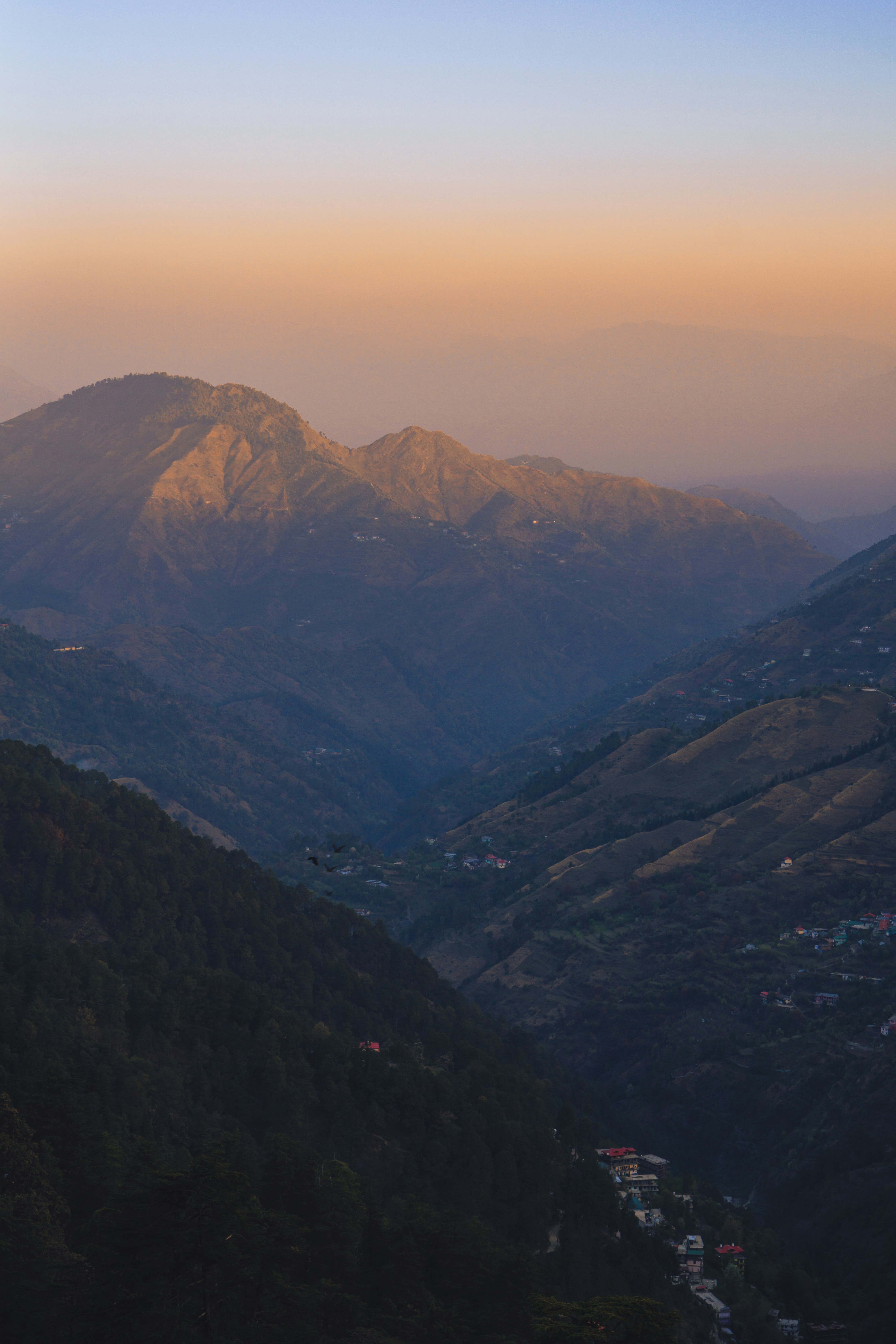 Golden-hued mountains under a soft twilight sky, revealing the serene beauty of a mountainous landscape. The valleys below are dotted with small settlements.