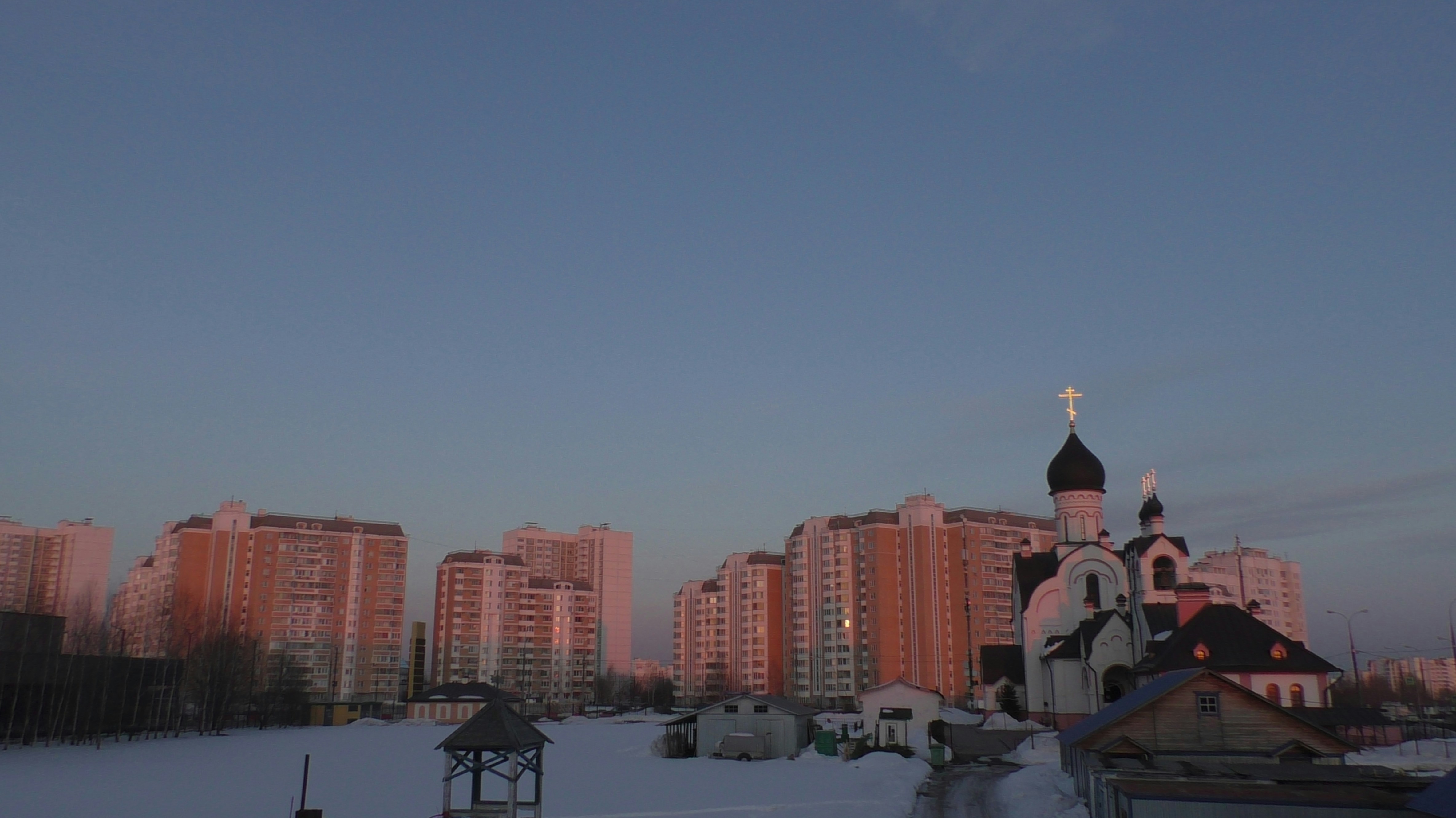 A serene winter scene featuring a quaint church juxtaposed against modern high-rise buildings, highlighting the contrast between tradition and contemporary life.