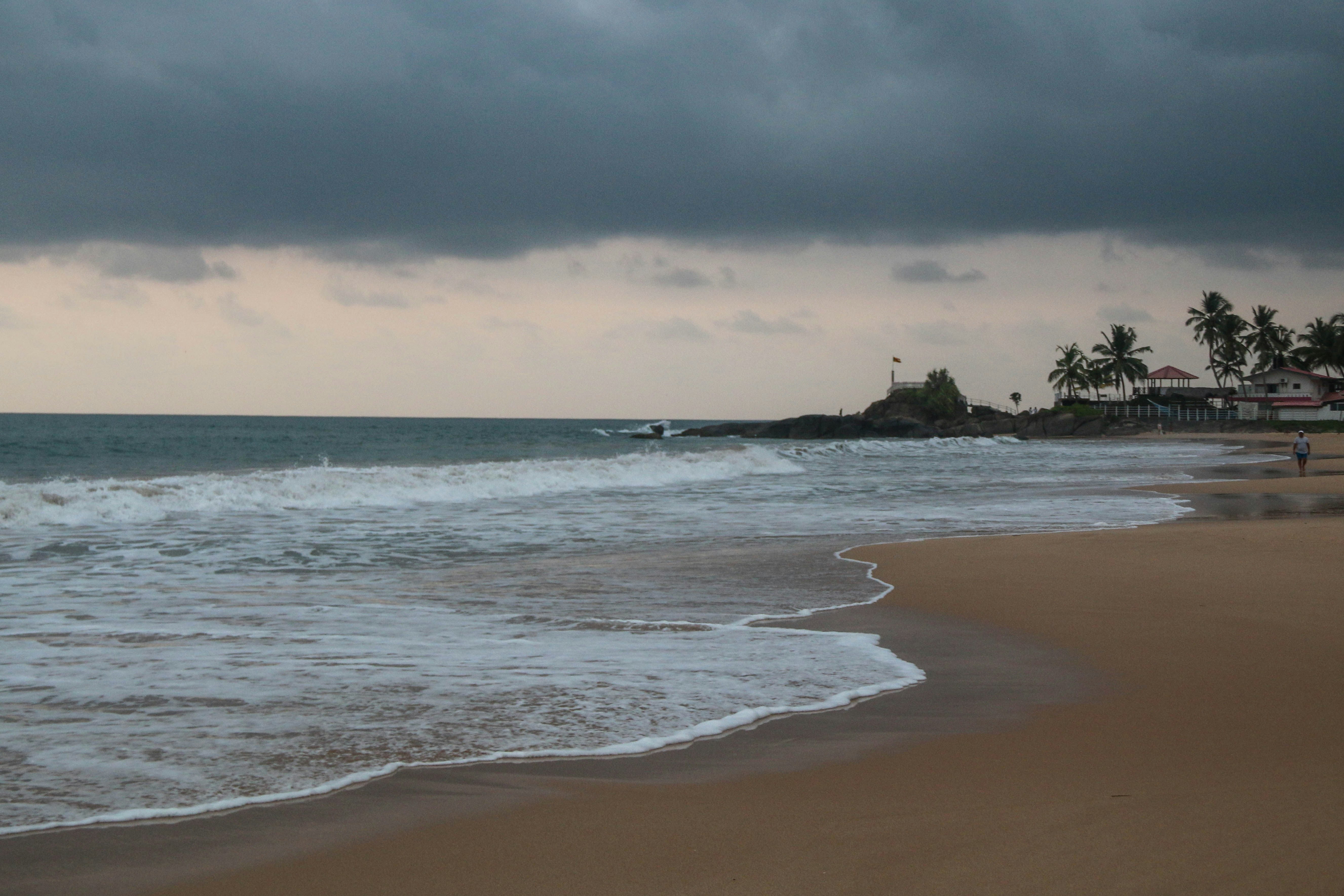 Waves gently lap against a sandy beach under a moody sky, with palm trees lining the shore and a distant structure on the rocks. The scene evokes a tranquil yet dramatic coastal atmosphere.