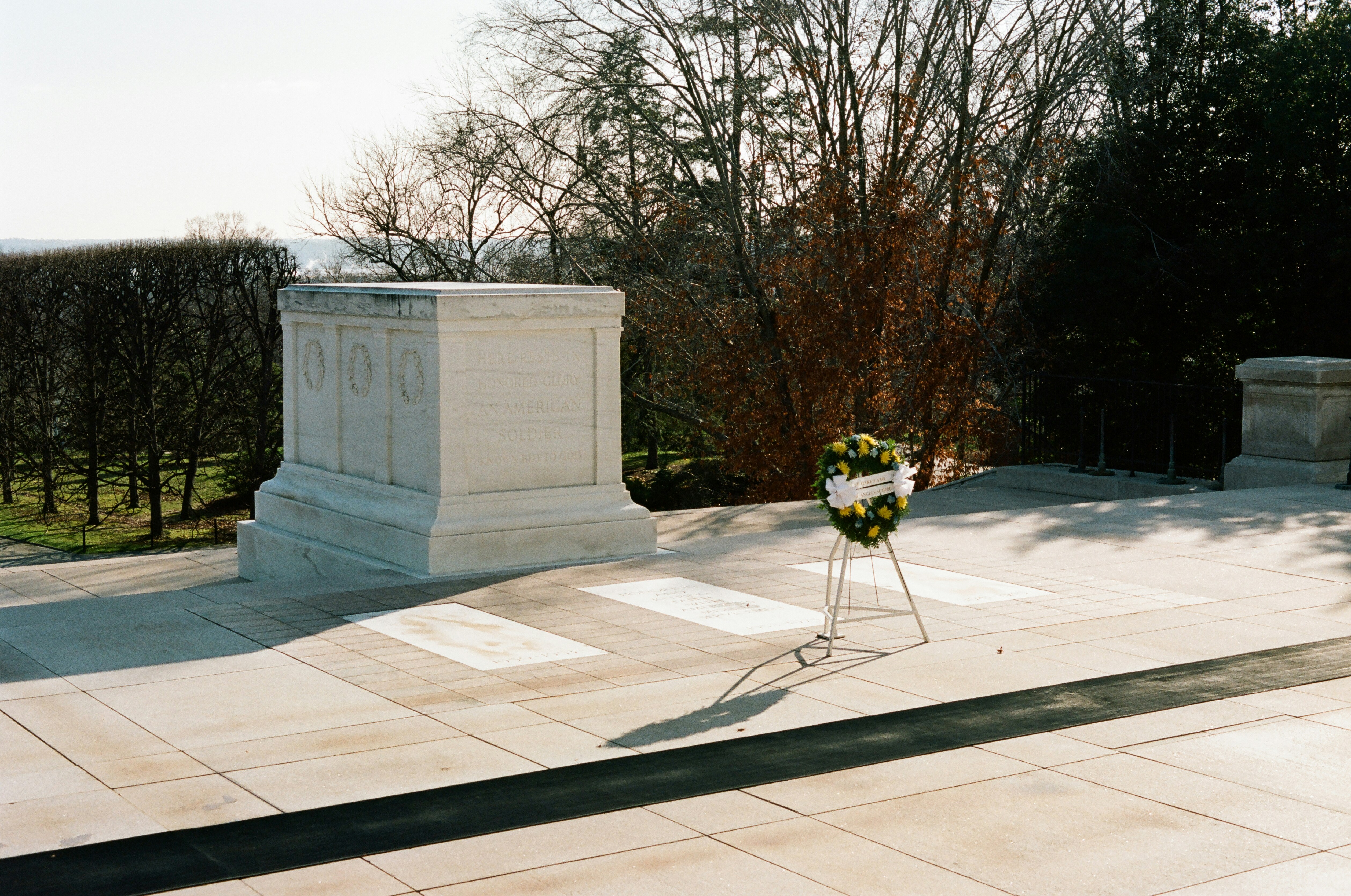 Stone memorial with a wreath in a sunlit park, framed by trees and a clear sky.