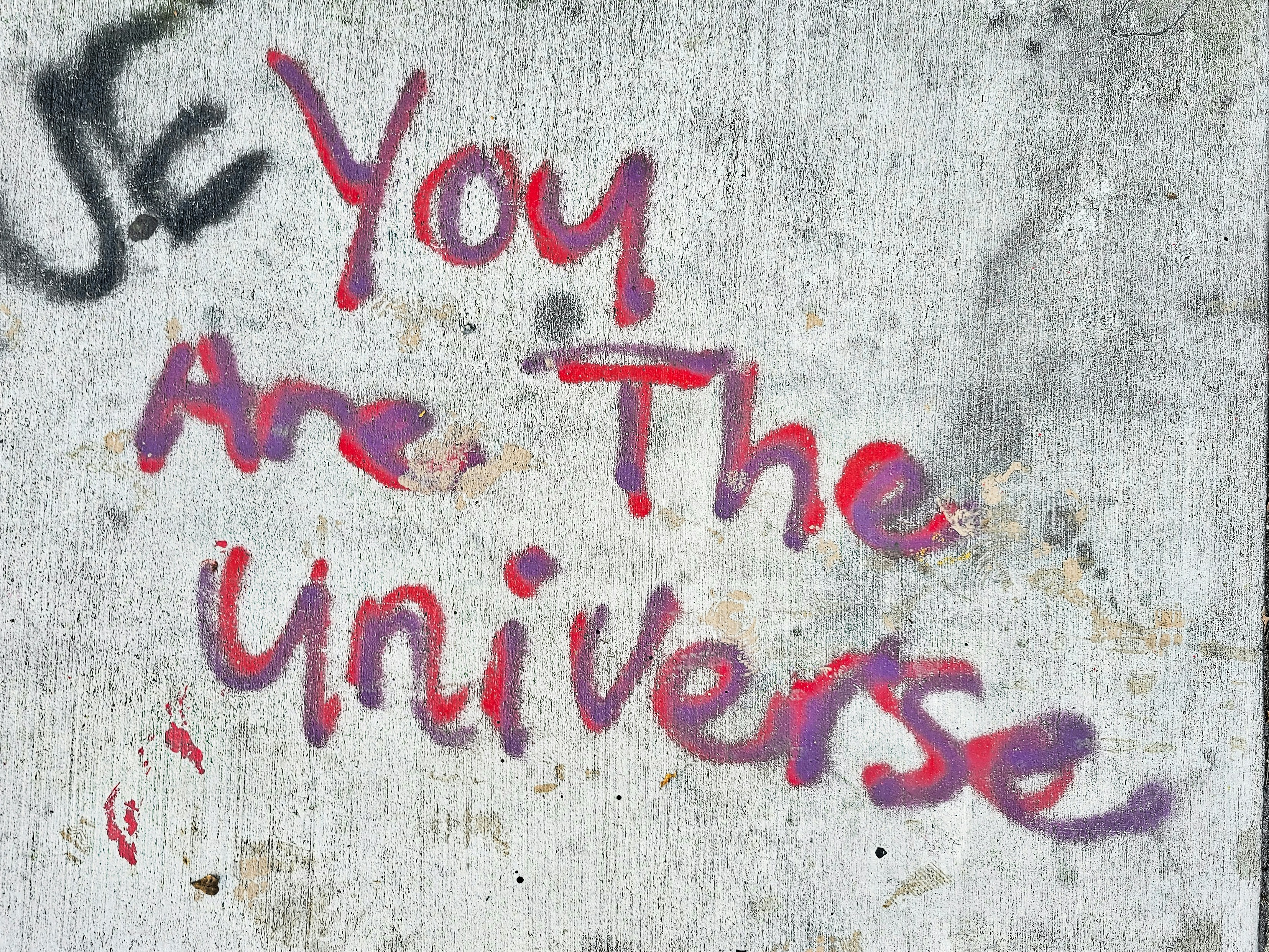Graffiti mural on a rough concrete wall reads 'You Are The Universe' in red and purple lettering.