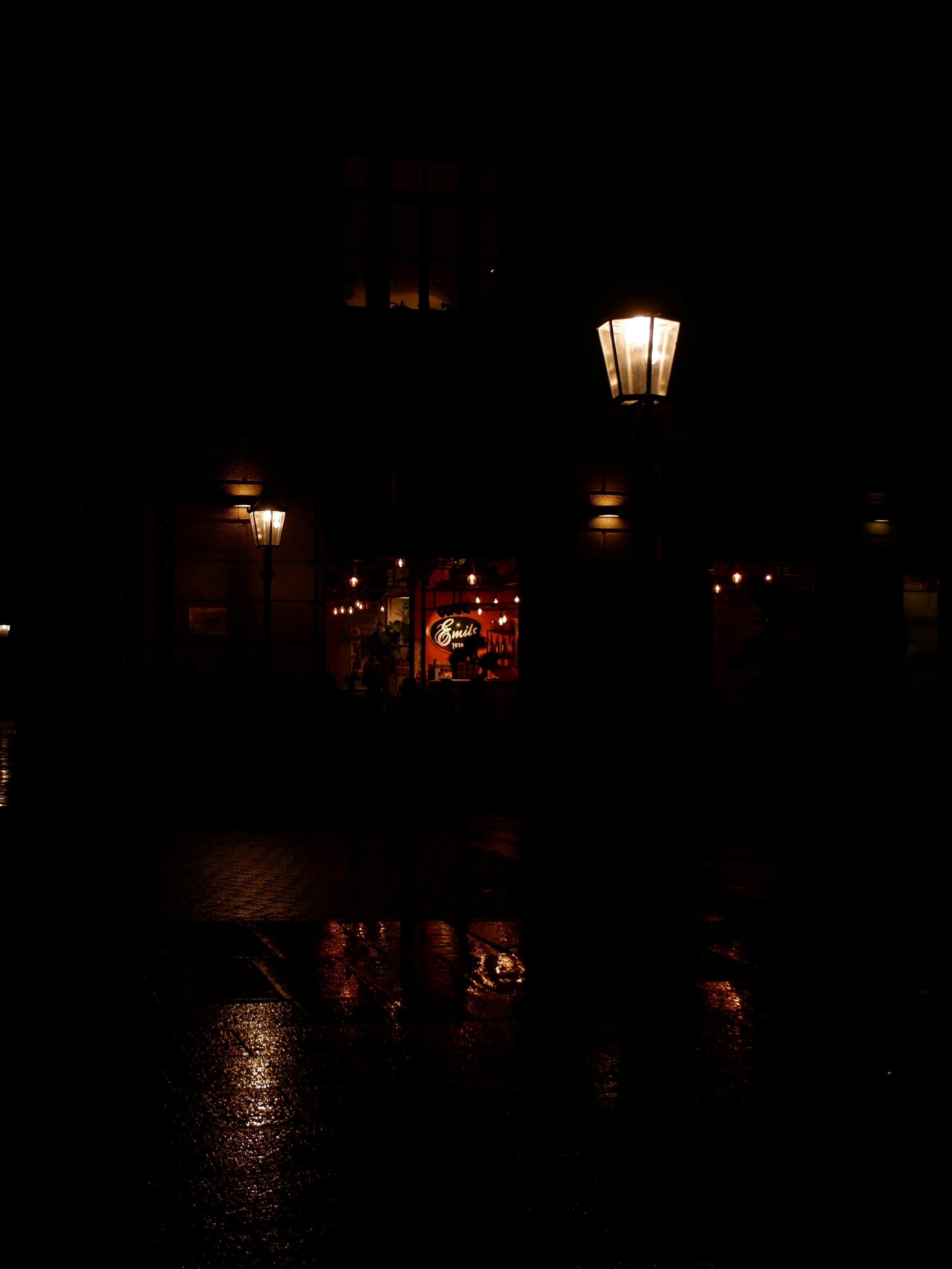 Nighttime photograph of a rain-washed square with warm street lamps and a brightly lit storefront window, reflections glistening on the wet pavement.