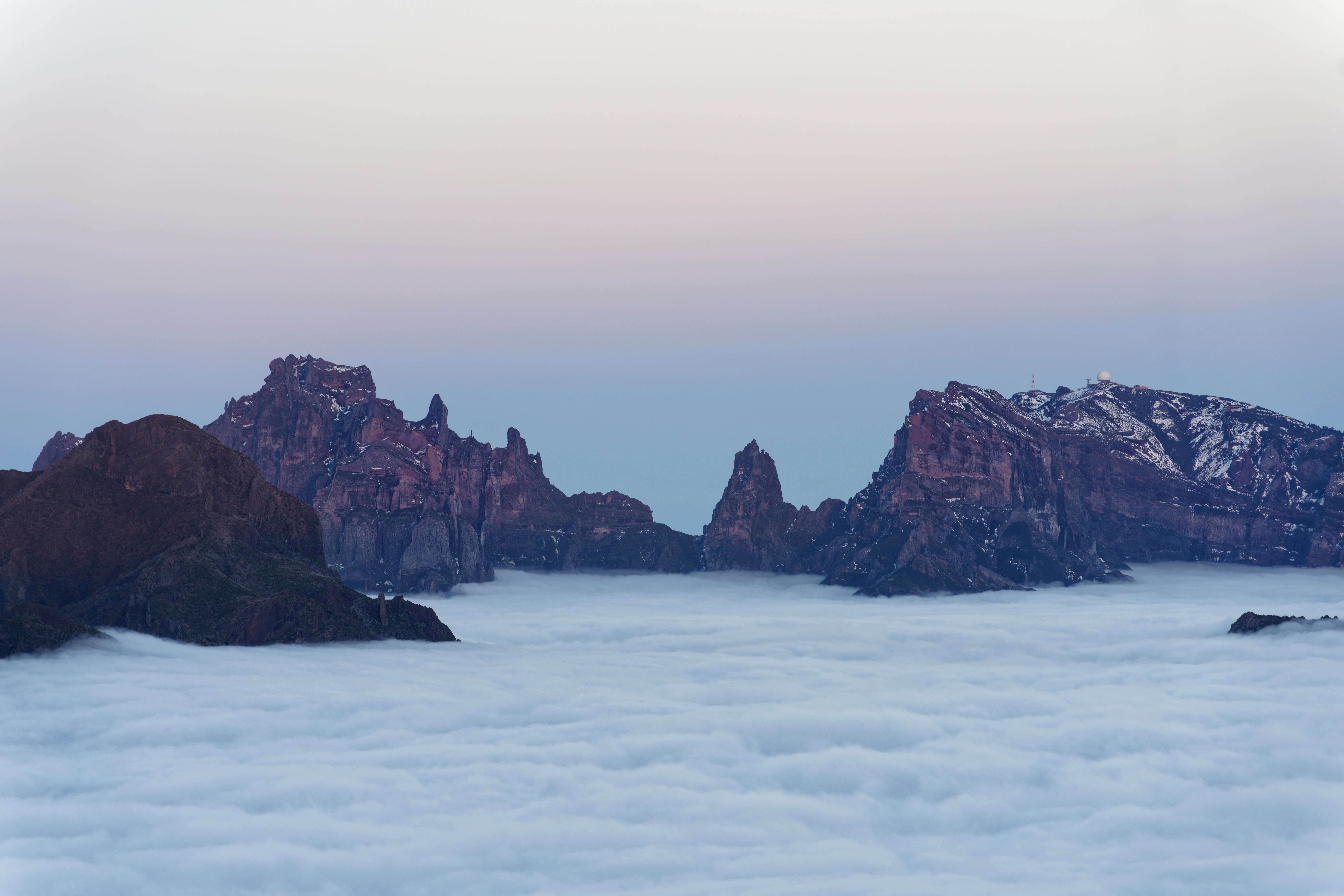 Jagged mountain silhouettes rise above a sea of clouds during twilight, creating a serene and mystical atmosphere.