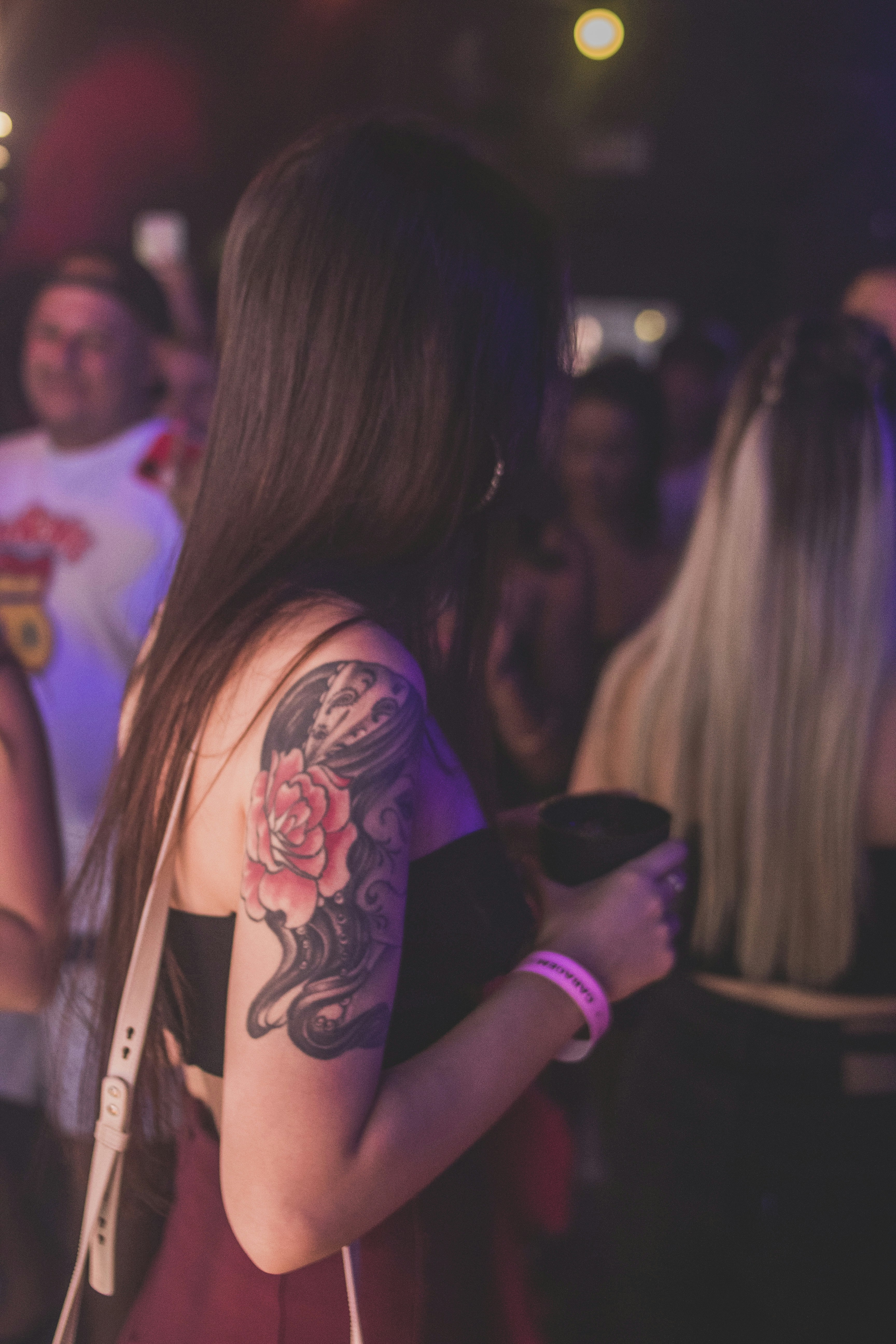 A woman with a floral tattoo enjoys a lively atmosphere, holding a drink amidst a crowd at a vibrant event.
