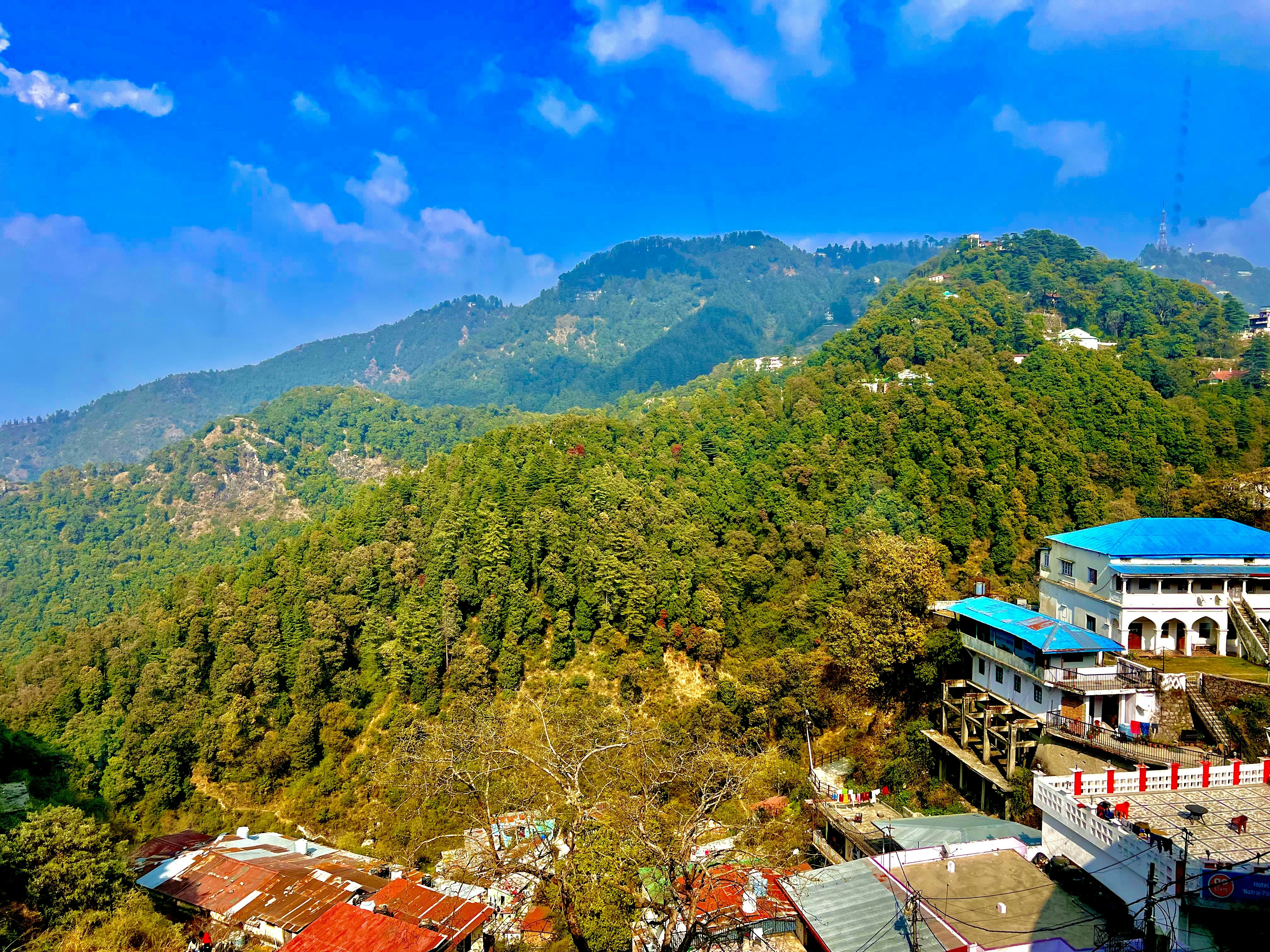 Lush green hills rise majestically against a bright blue sky, with quaint buildings nestled at the base. The scene captures the harmony between nature and human habitation.