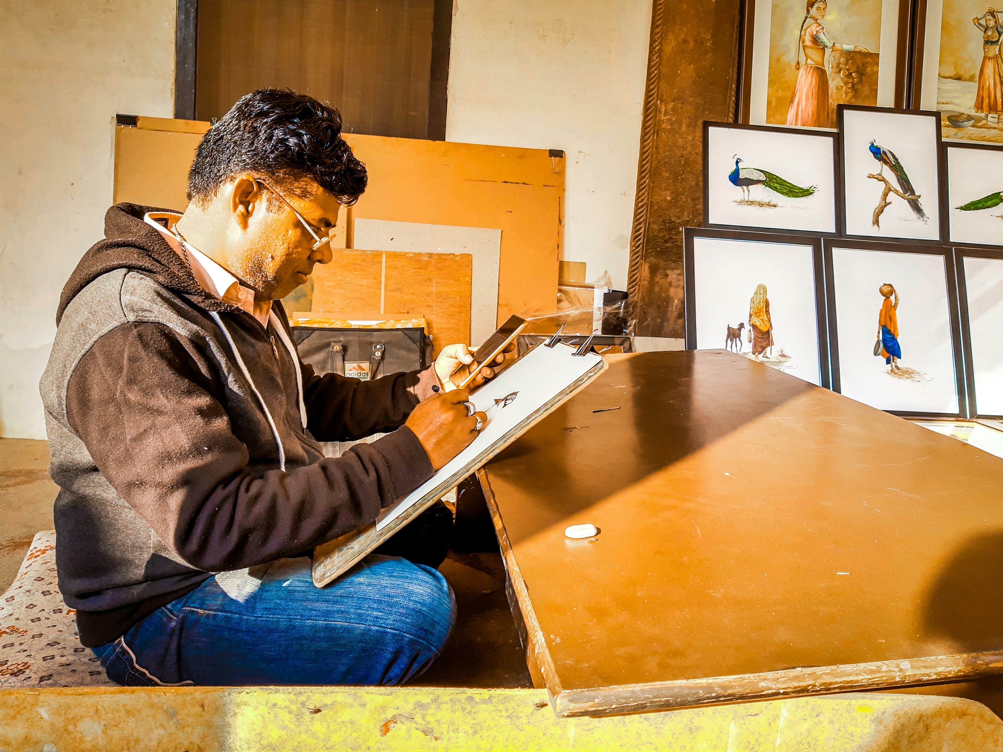 Artist engrossed in sketching at a wooden table, surrounded by framed illustrations in a warmly lit room.
