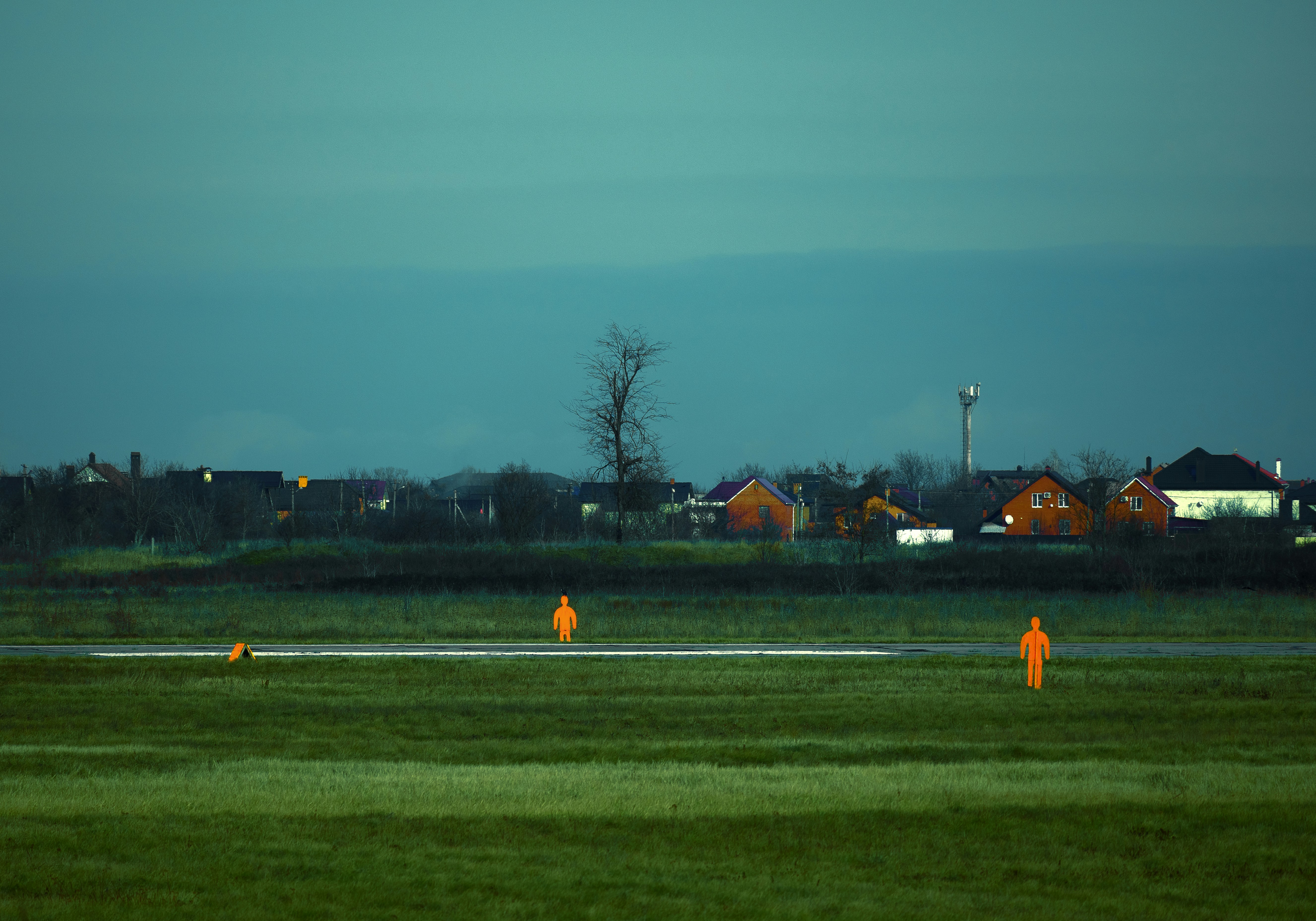 Two figures dressed in orange stand on a grassy field, contrasting against a distant backdrop of houses and a solitary tree. The scene evokes a sense of isolation and contemplation.