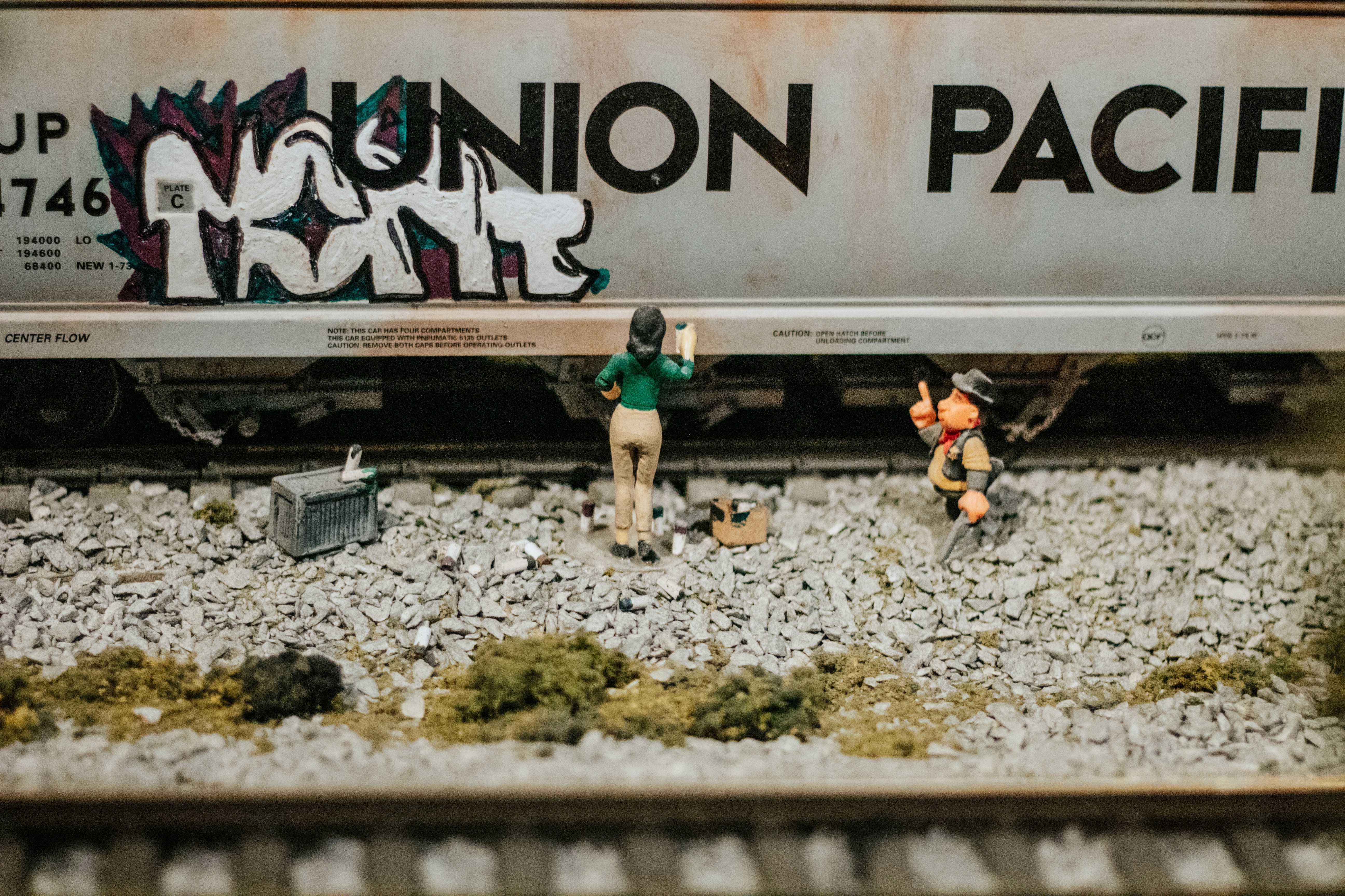 Tiny Figures at the Tracks: A Micro Scene Beneath a Graffiti-Streaked Freight Car