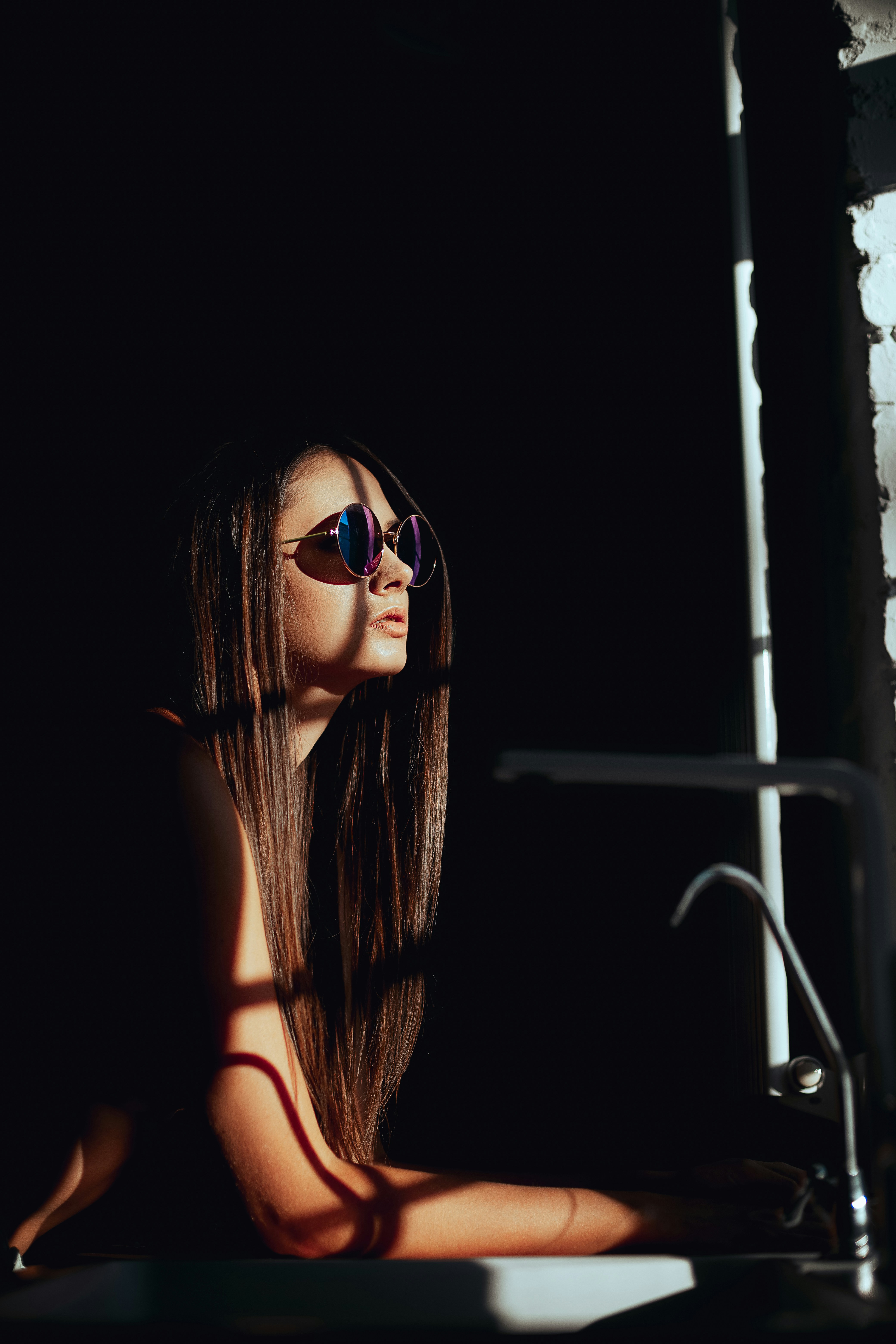 A woman with long hair and round sunglasses gazes thoughtfully, illuminated by soft light streaming through a window, creating a striking contrast with the dark background.