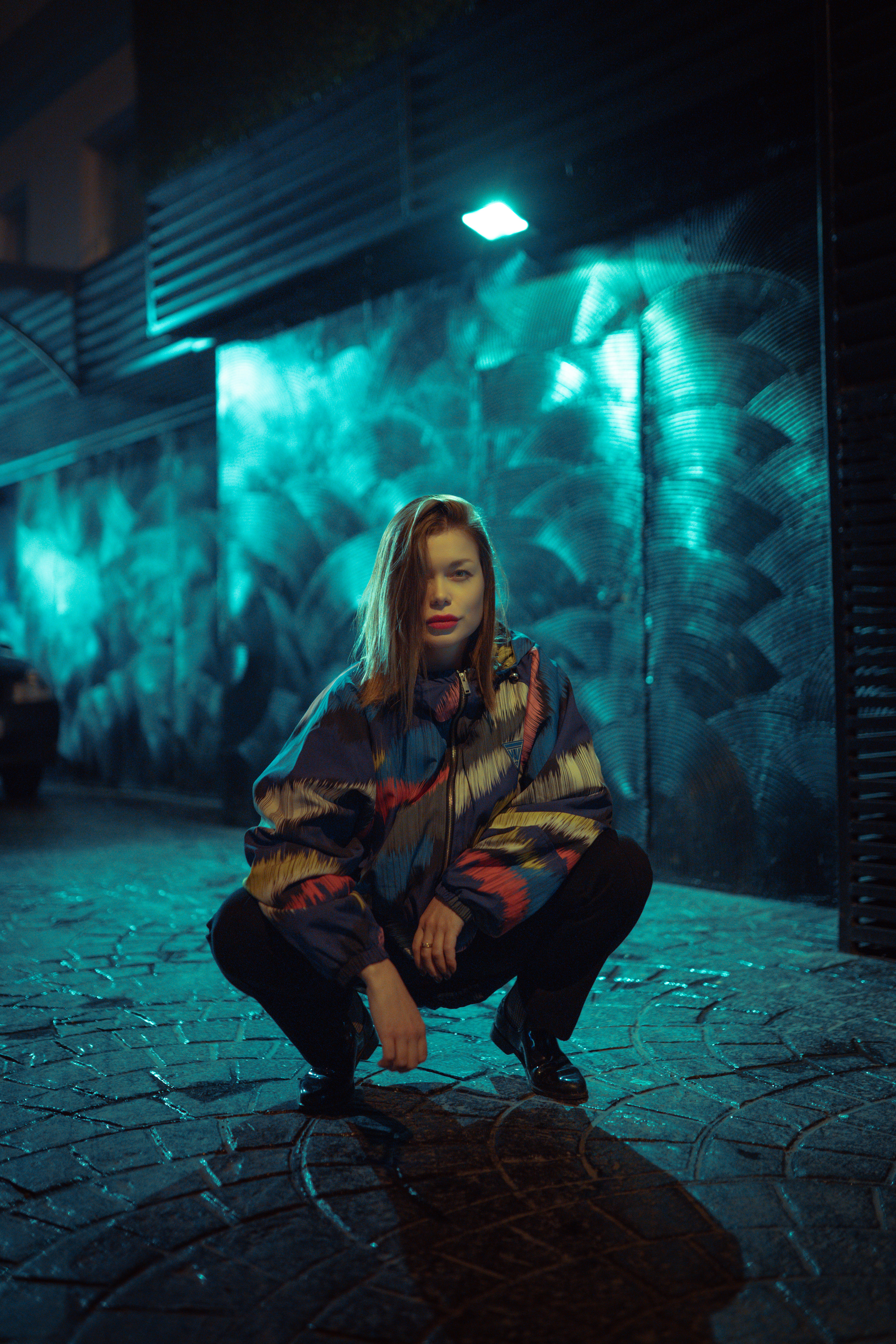 A woman squats in a vibrant, oversized jacket against a backdrop of illuminated graffiti in a nighttime urban setting.