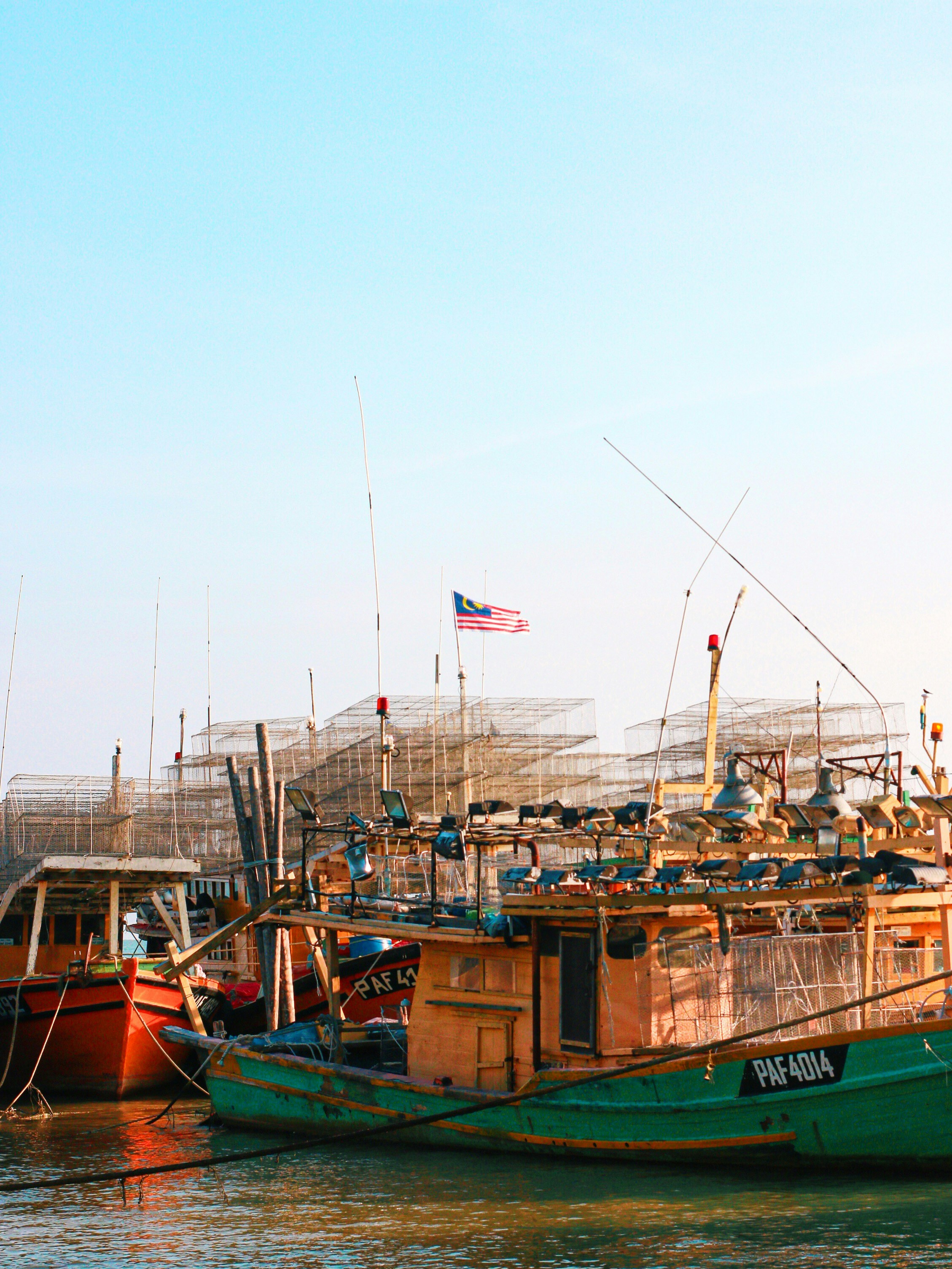 Colorful fishing boats moored in a tranquil harbor with a gentle breeze, featuring a Malaysian flag waving in the background.
