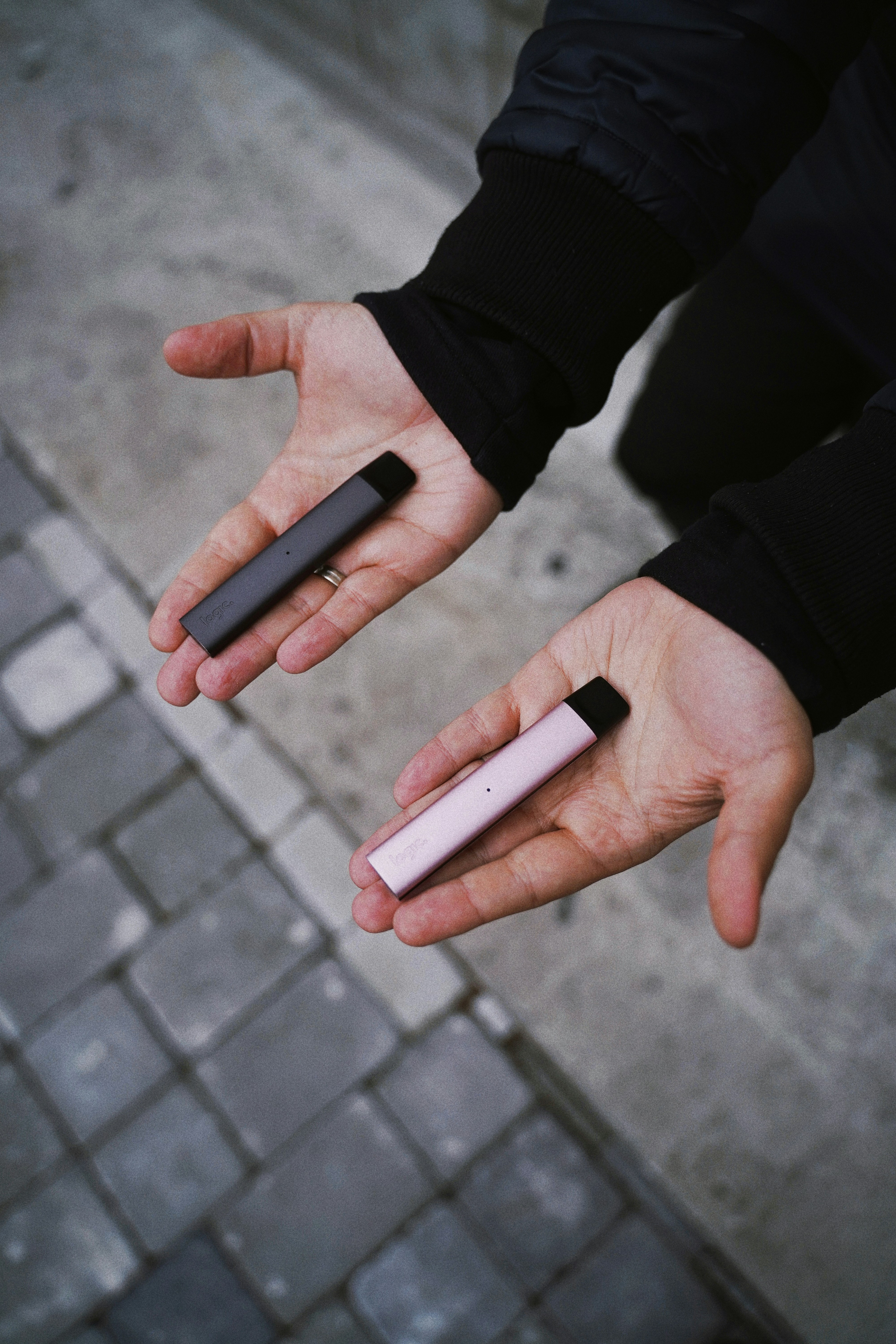 Two vaping devices held in open hands, one black and one pink, against a textured background. The scene highlights the modern trend of vaping.