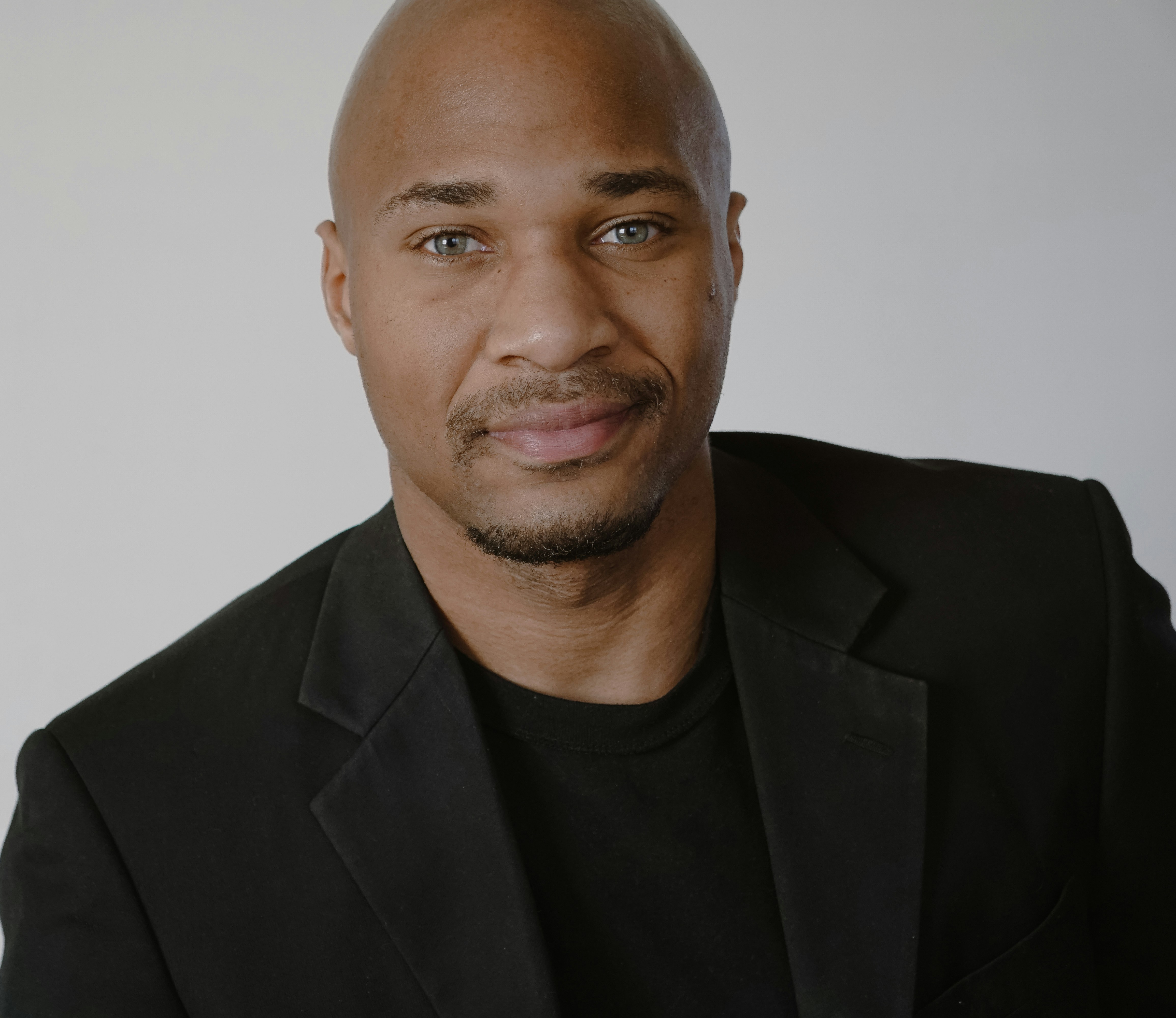 A man in a black blazer and black shirt poses confidently against a soft, neutral background.