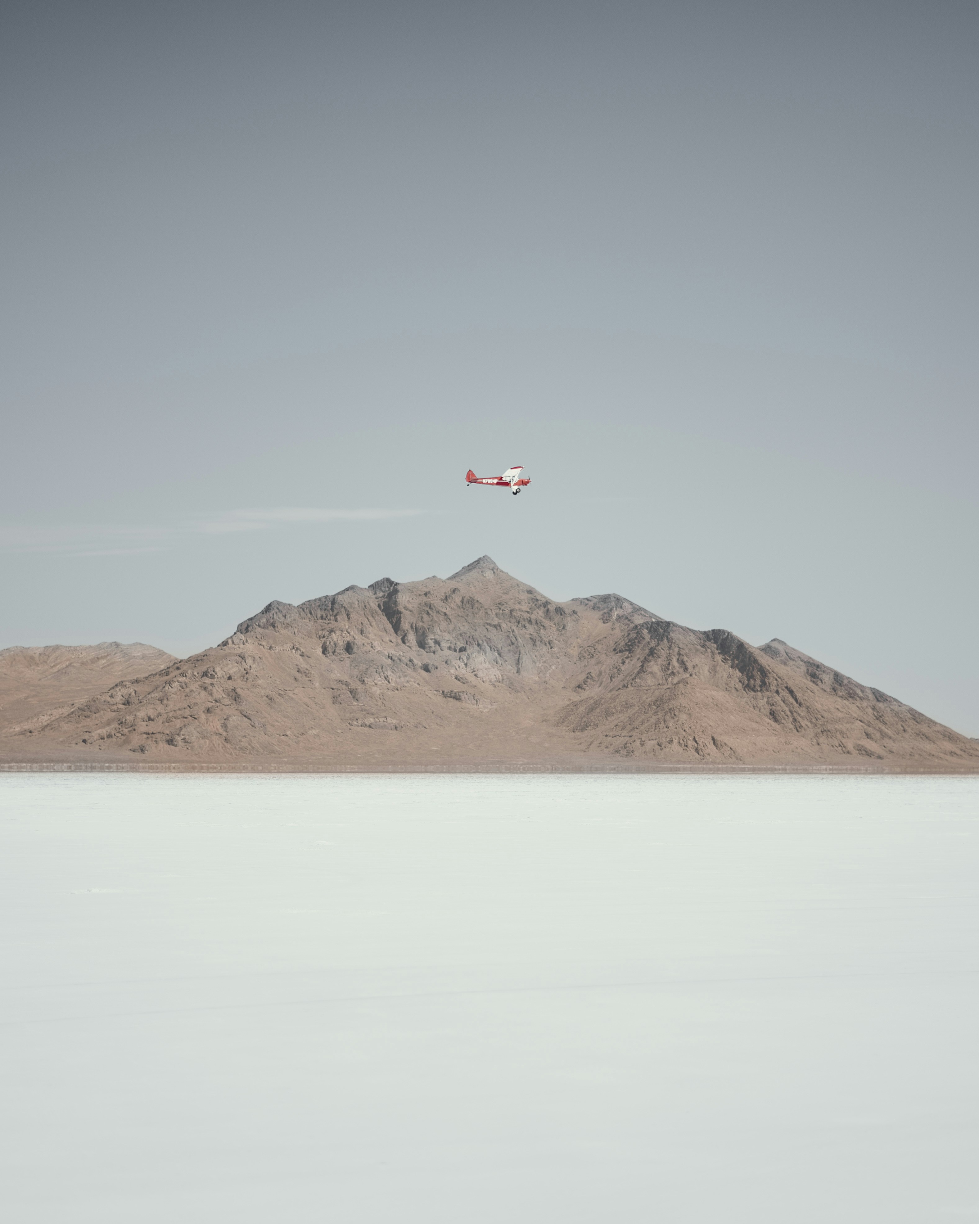 A small red plane glides gracefully over expansive salt flats, with distant mountains providing a stark backdrop.