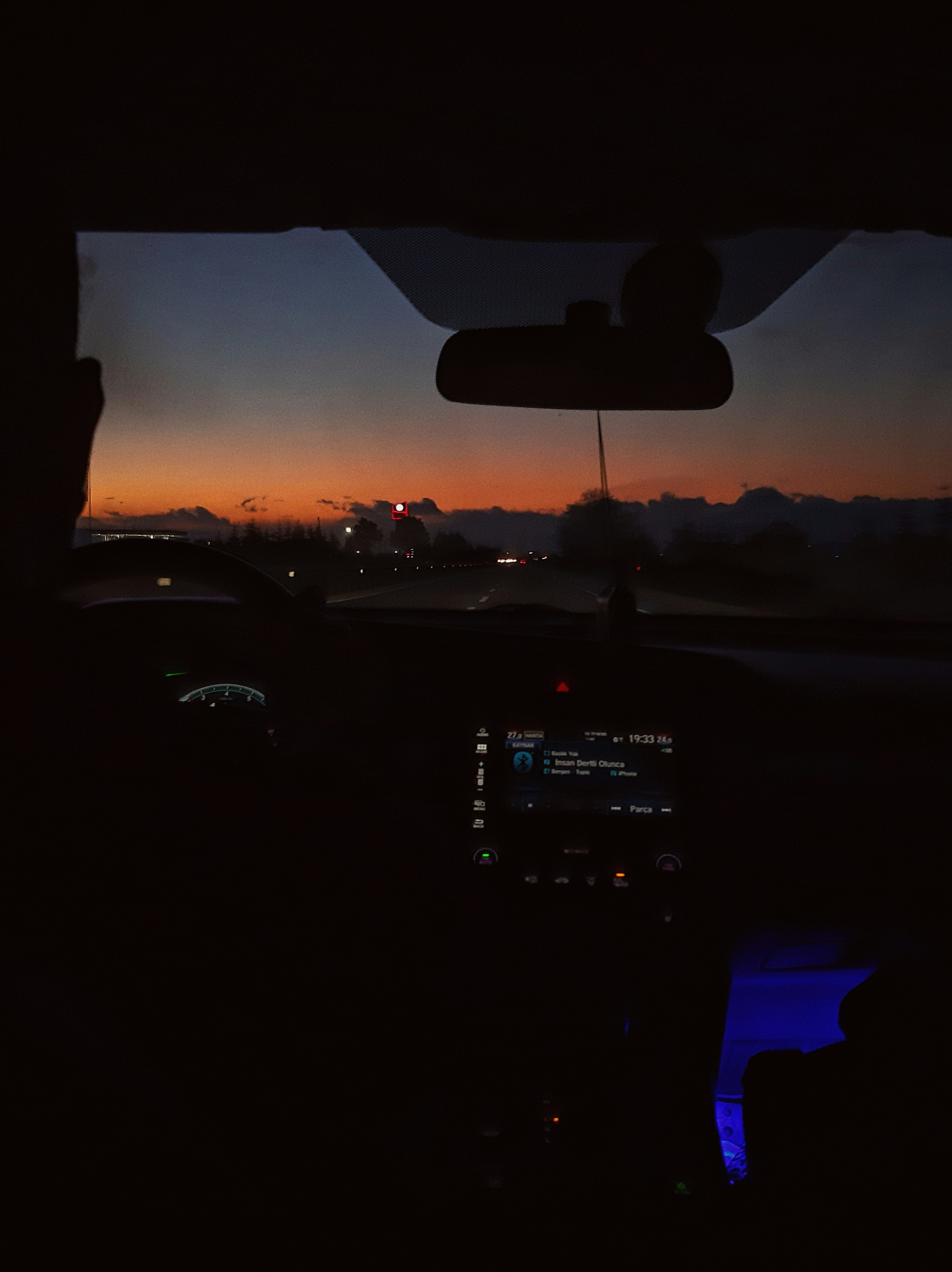Silhouette of a driver in a car against a vibrant twilight sky, with dashboard lights illuminating the interior. 