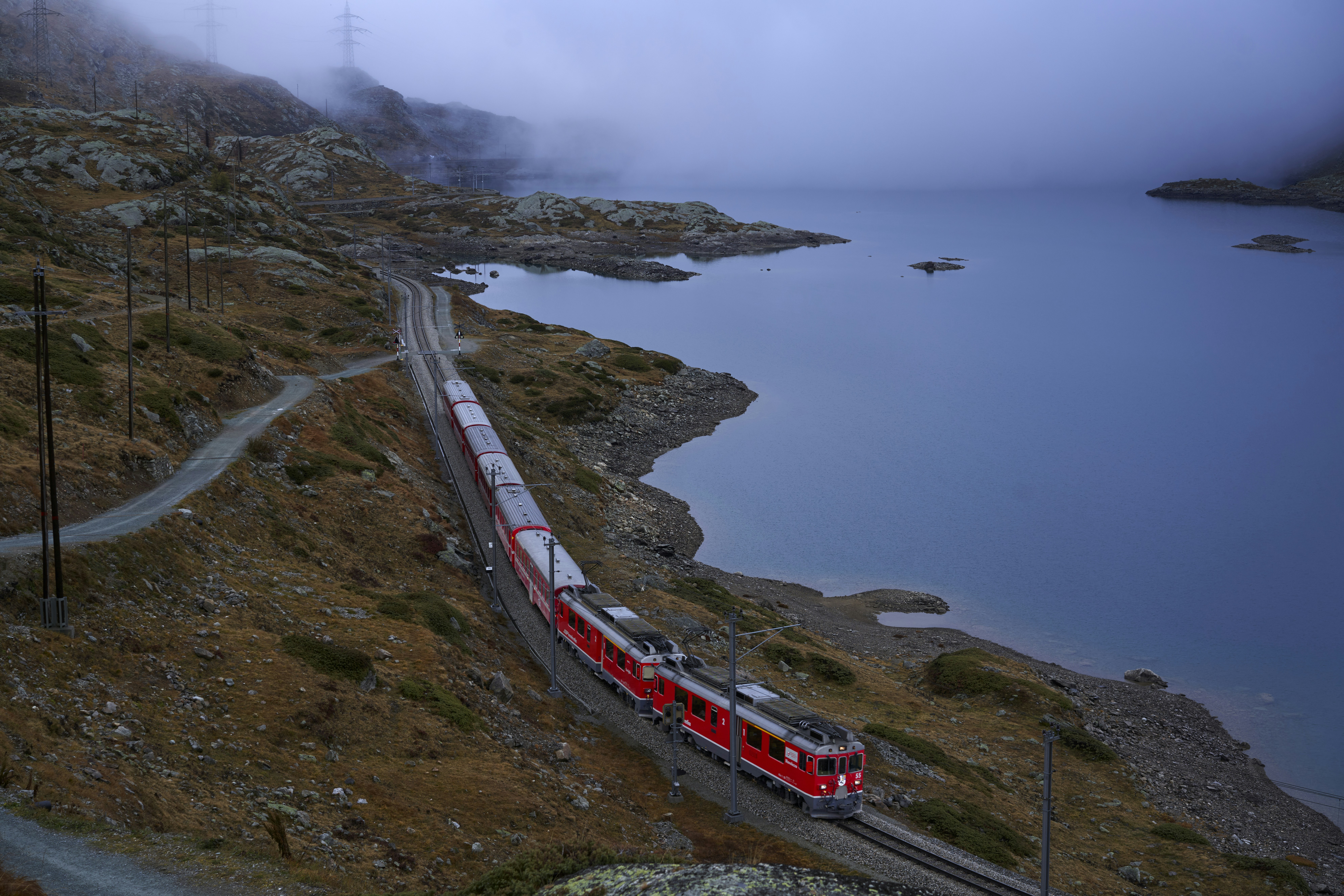 Red train winding along a rugged lakeside track, enveloped in thick fog and rocky terrain.