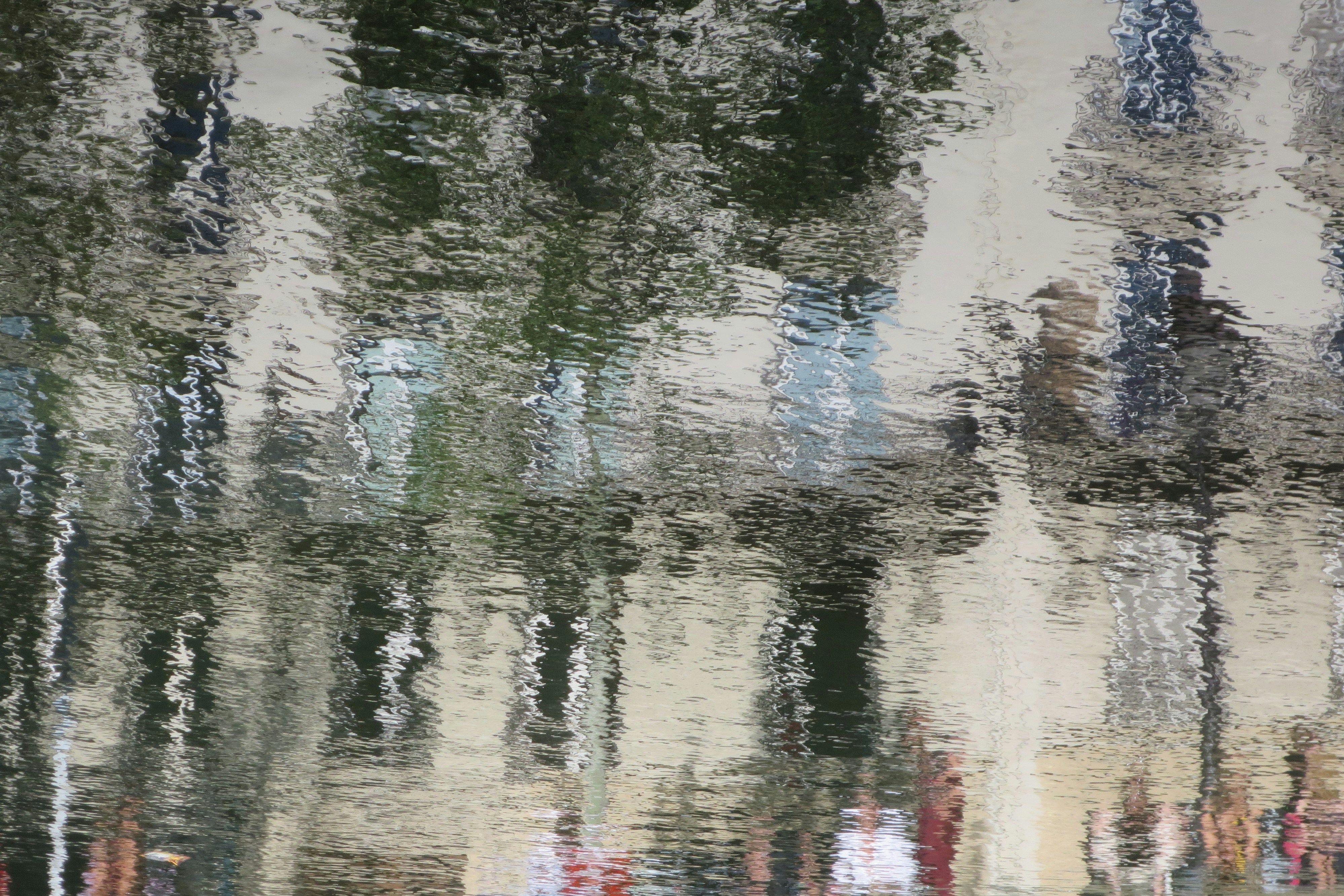 Distorted reflections of buildings and people ripple across a water surface, creating an abstract interplay of colors and shapes.
