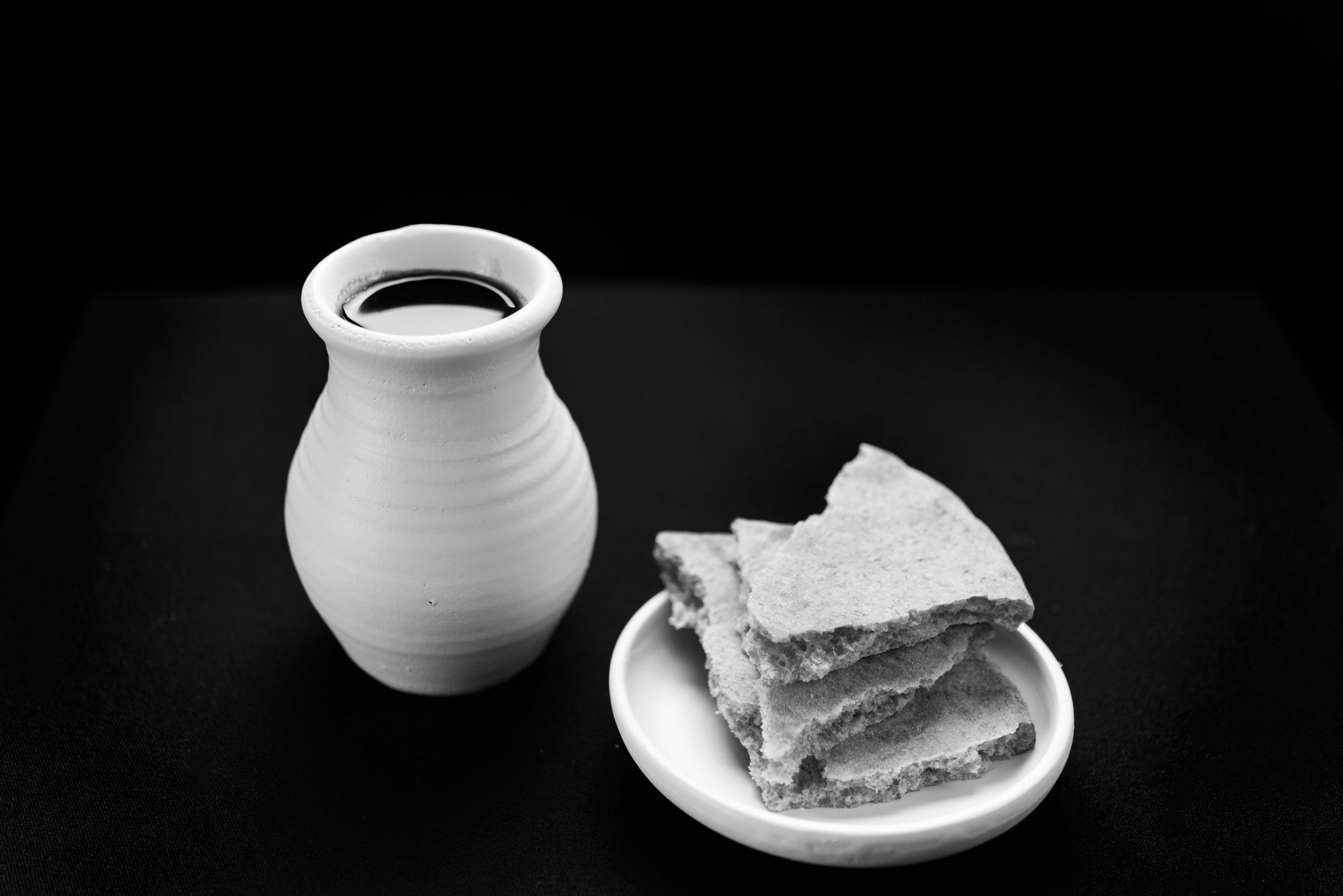 Ceramic jug filled with olive oil next to a plate of rustic bread slices on a black background.