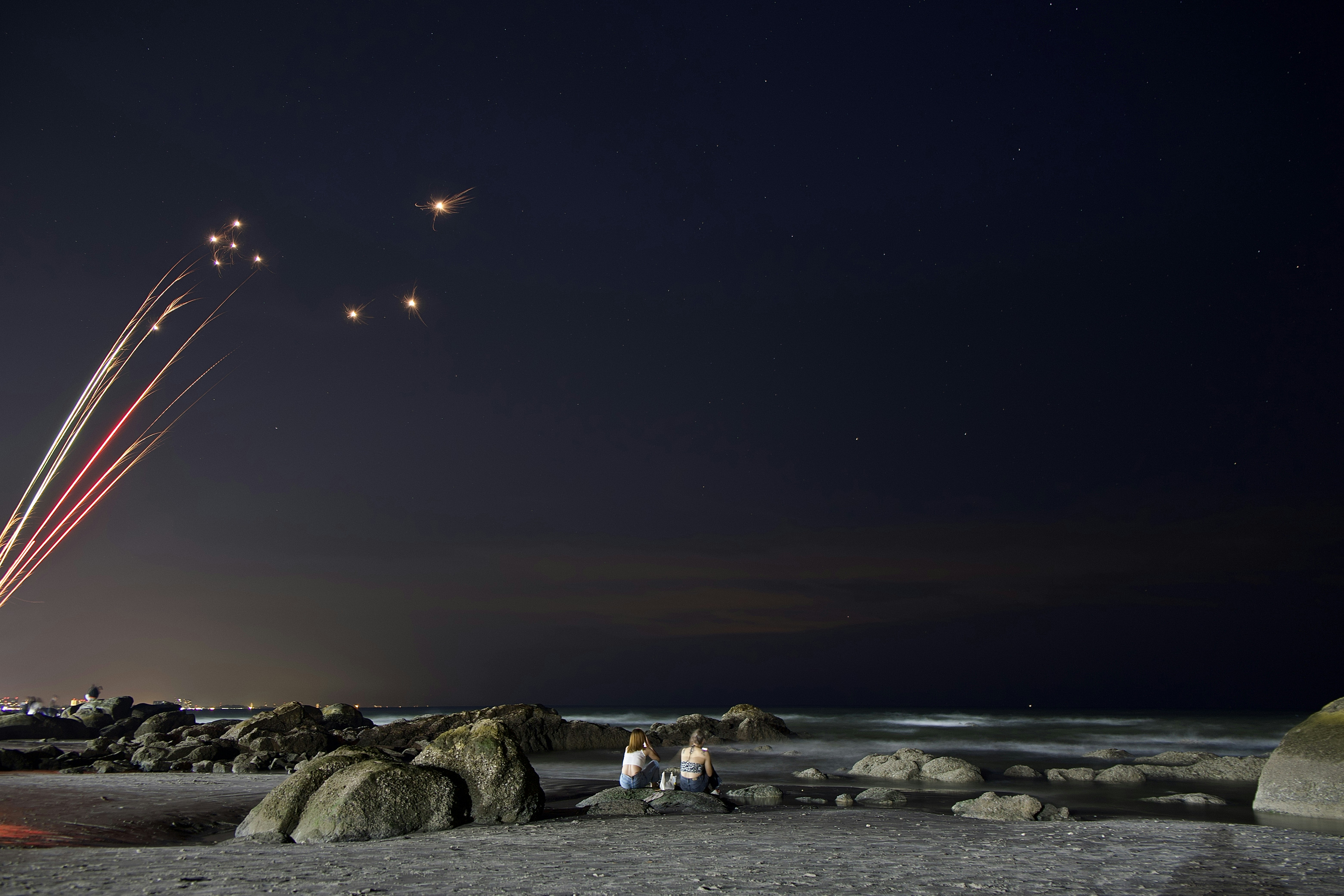 Two figures seated on a rocky beach, gazing at colorful aerial fireworks against a starry night sky.