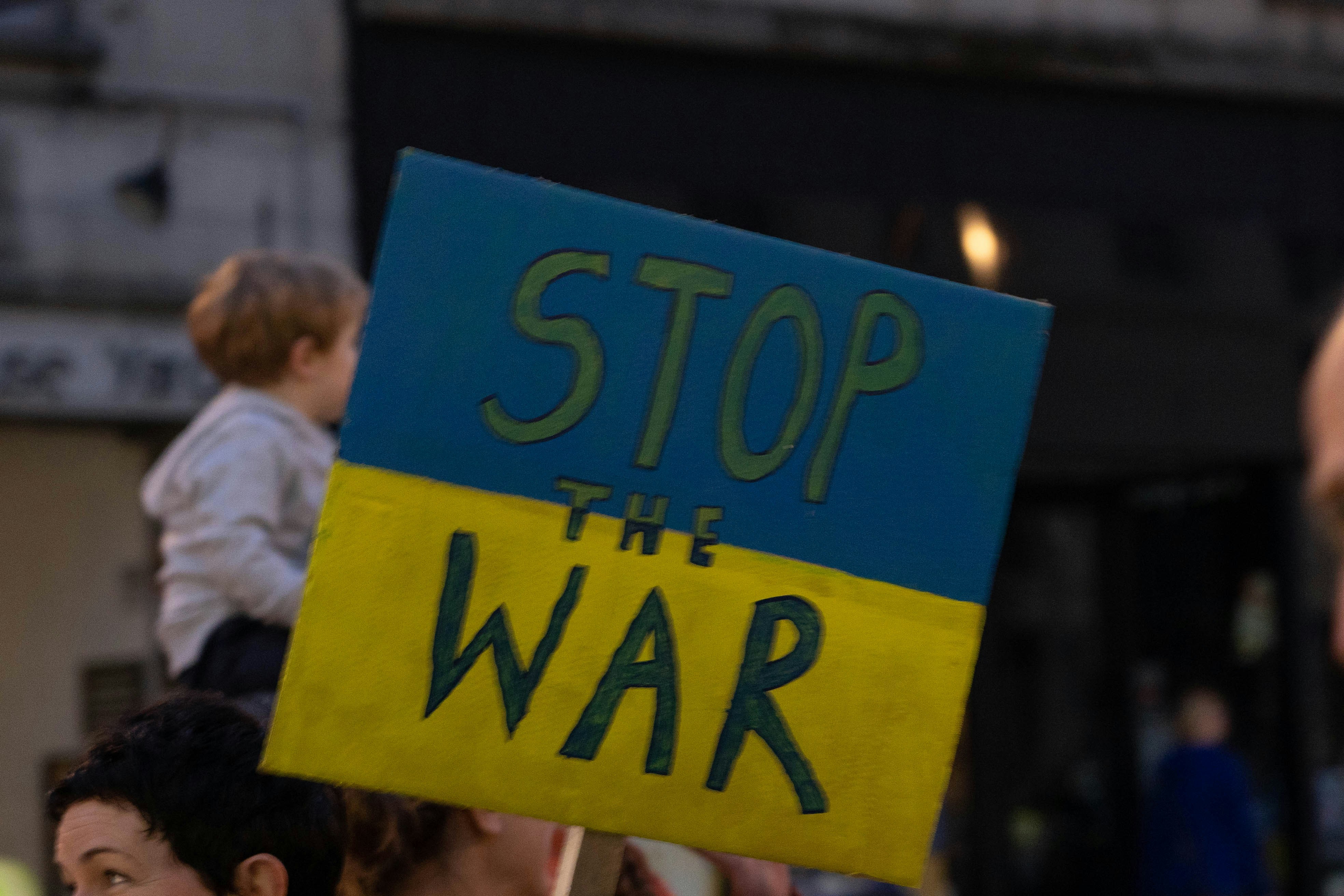 Vibrant protest sign reading 'STOP THE WAR' in blue and yellow, held by a demonstrator with a child on their back. The backdrop suggests a lively public gathering.