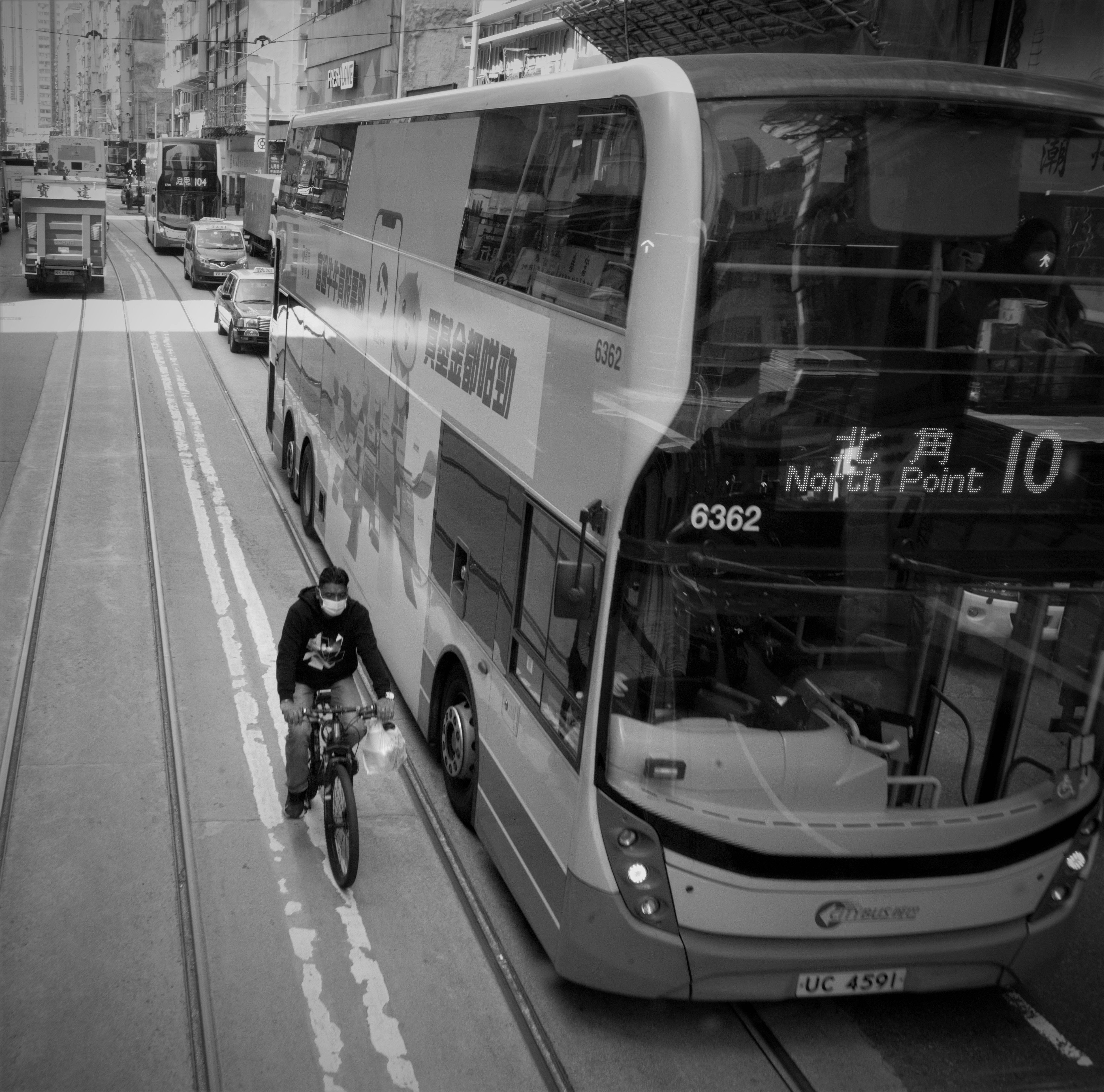 A cyclist navigates the bustling streets beside a double-decker bus in an urban landscape, showcasing the rhythm of city life.