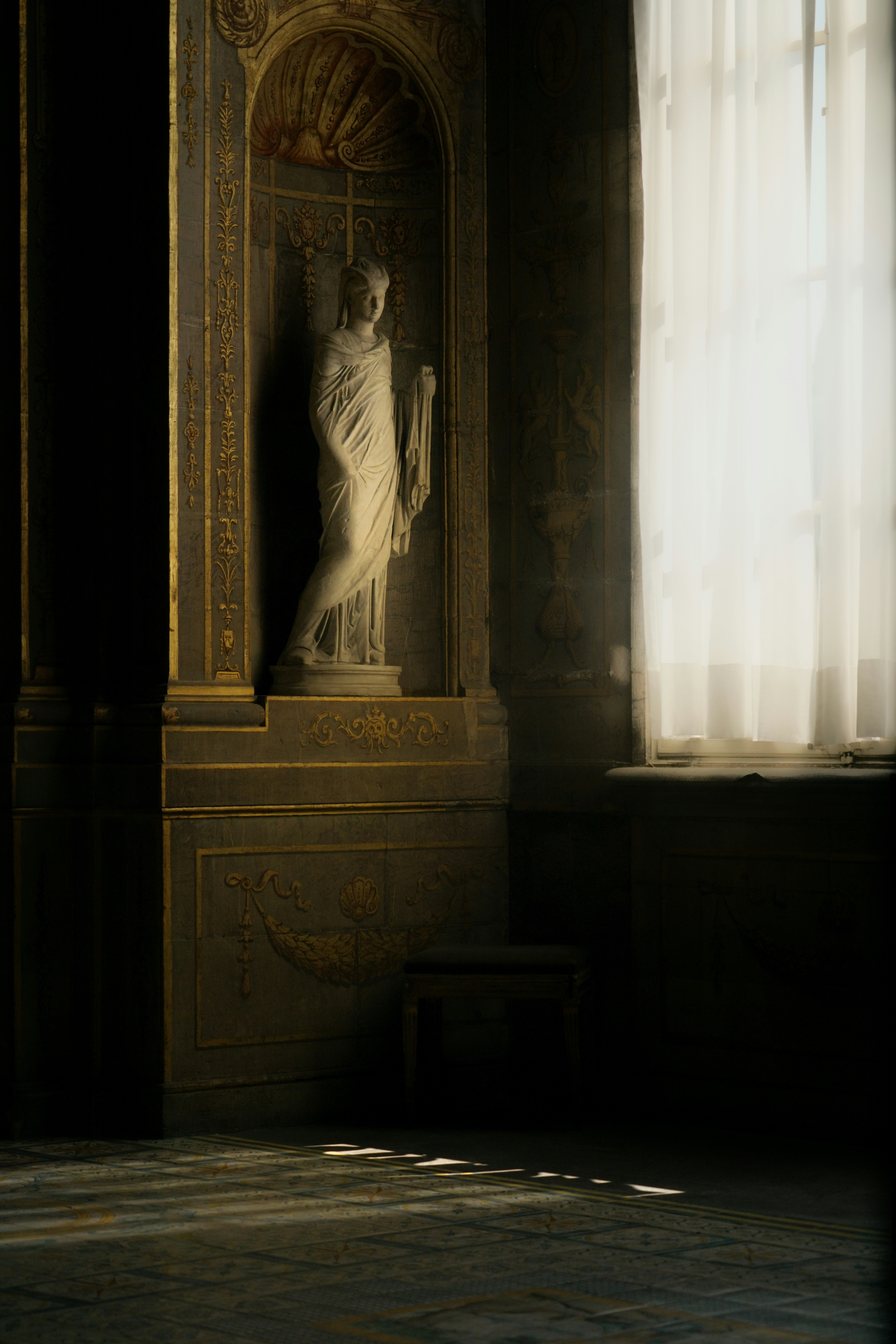 A marble statue of a figure draped in fabric stands in a softly lit corner, framed by intricate wall designs and a sheer curtain. The interplay of light creates a serene atmosphere.