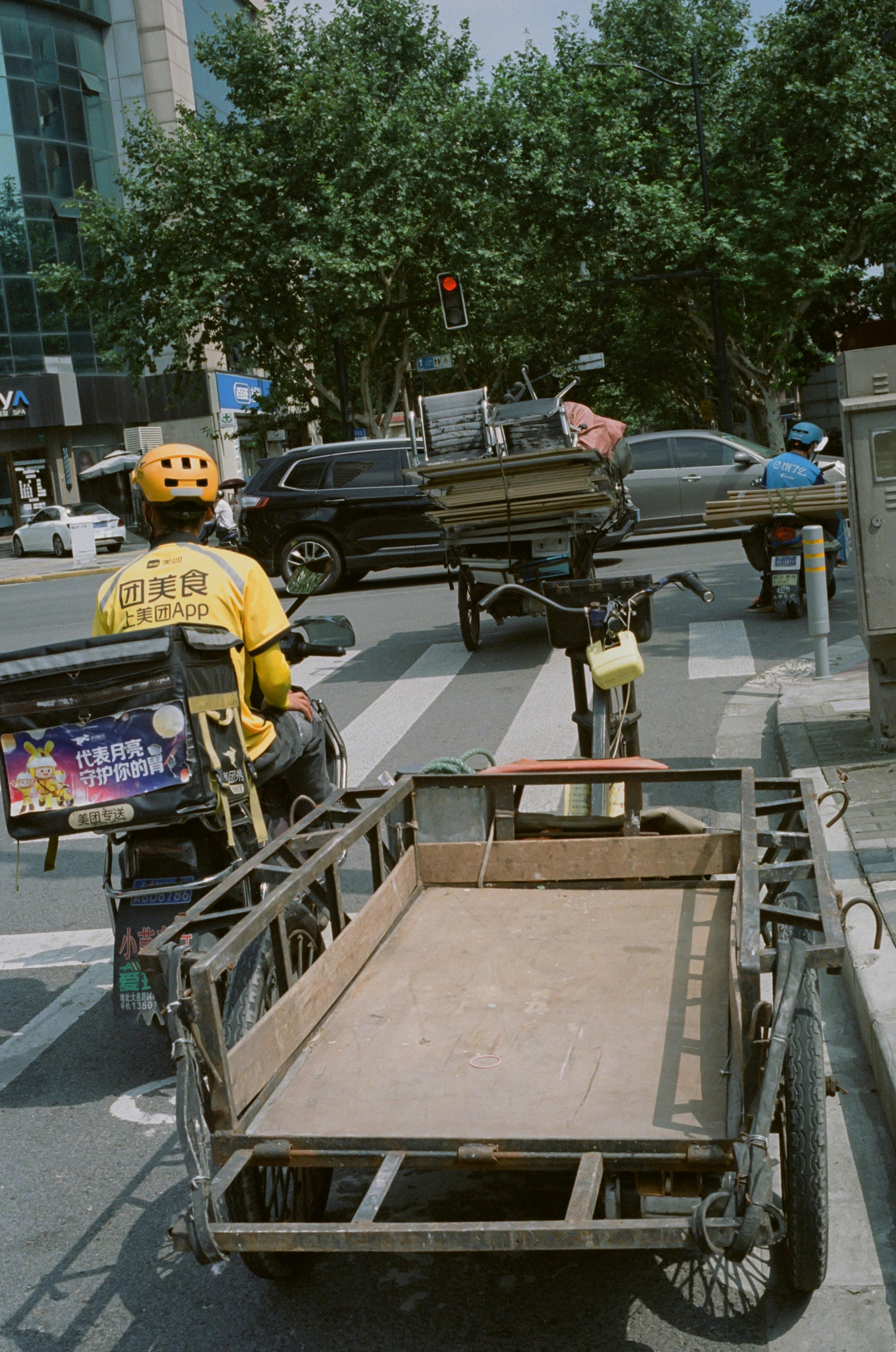 City street photograph of a delivery worker in a bright yellow jacket and helmet beside a flatbed cart, with a red traffic light overhead as cars pass.