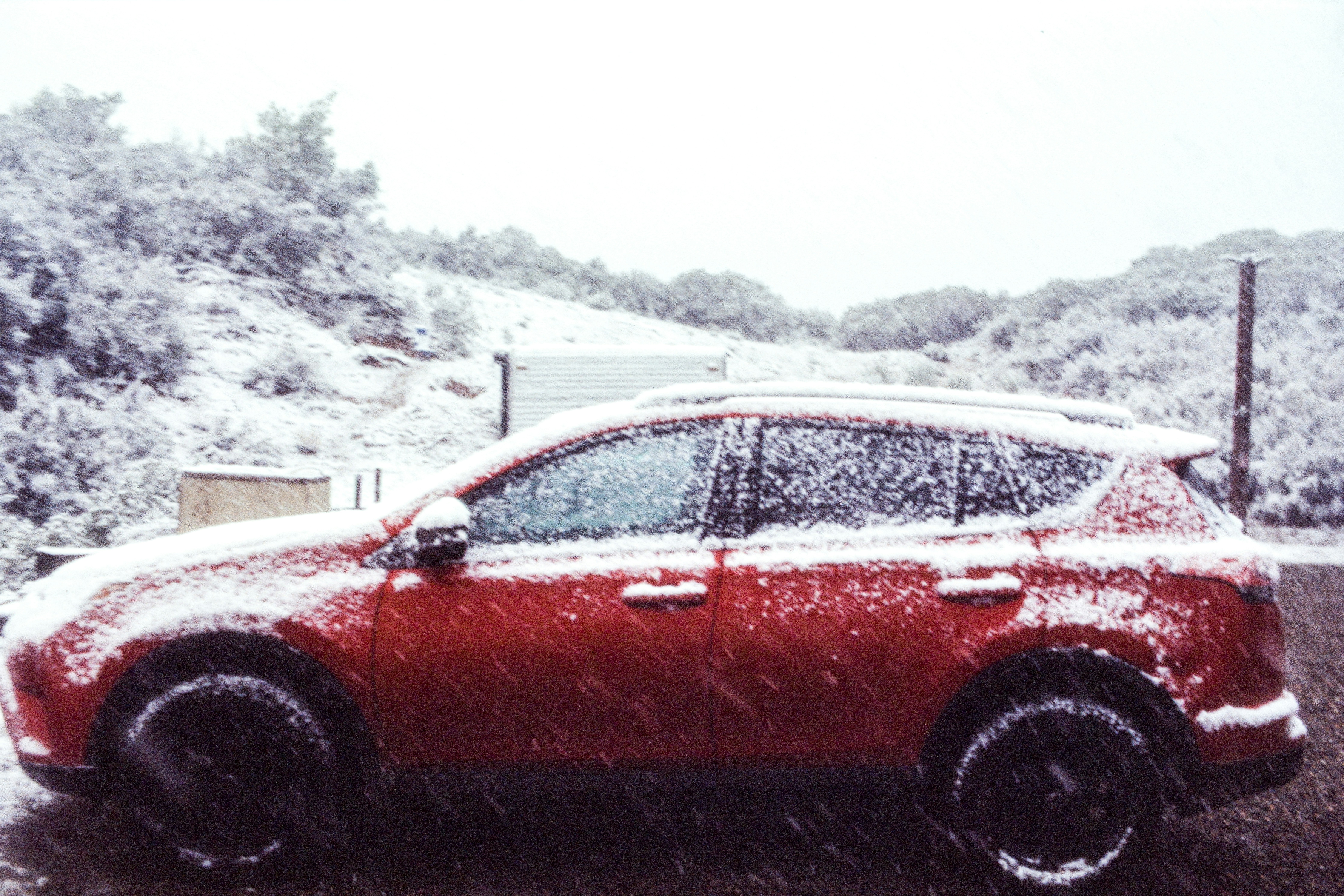 Red SUV parked in a snowy landscape, surrounded by a serene winter scene. Snowflakes gently accumulate on the vehicle's surface.