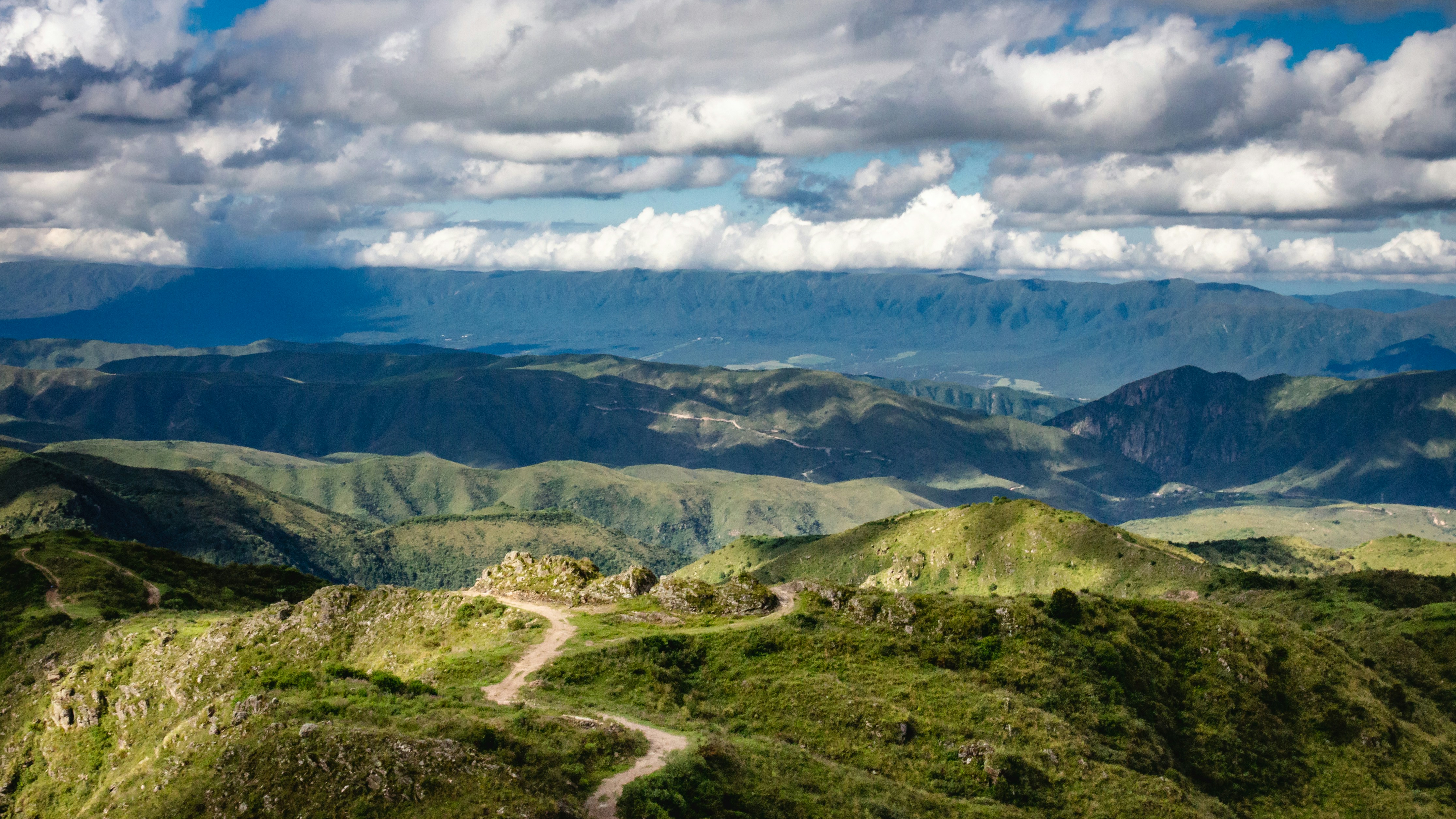 Mountain landscape with a winding path cutting through lush green hills under a sky filled with dramatic clouds.