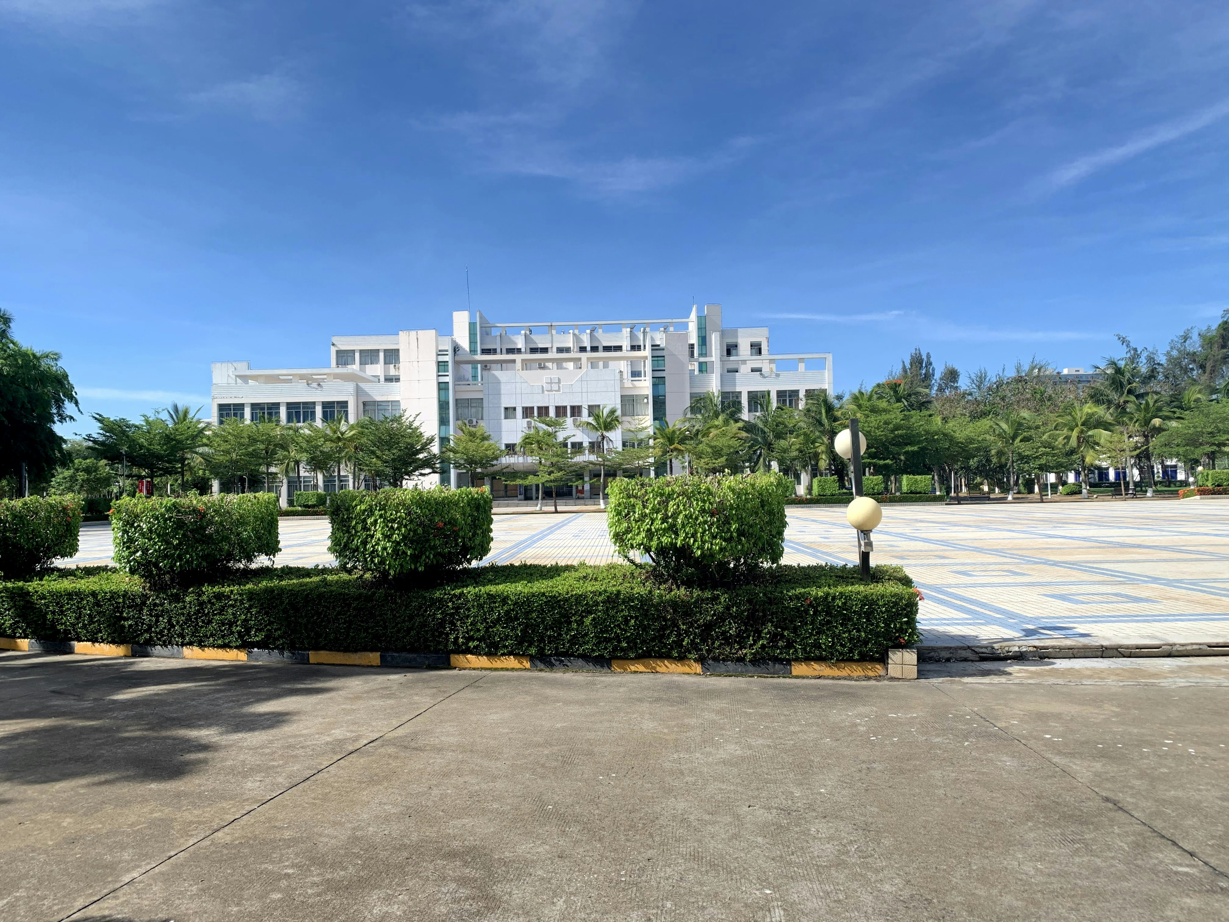 A contemporary building framed by manicured hedges and palm trees, set against a clear blue sky. The scene captures a tranquil outdoor space designed for gatherings.