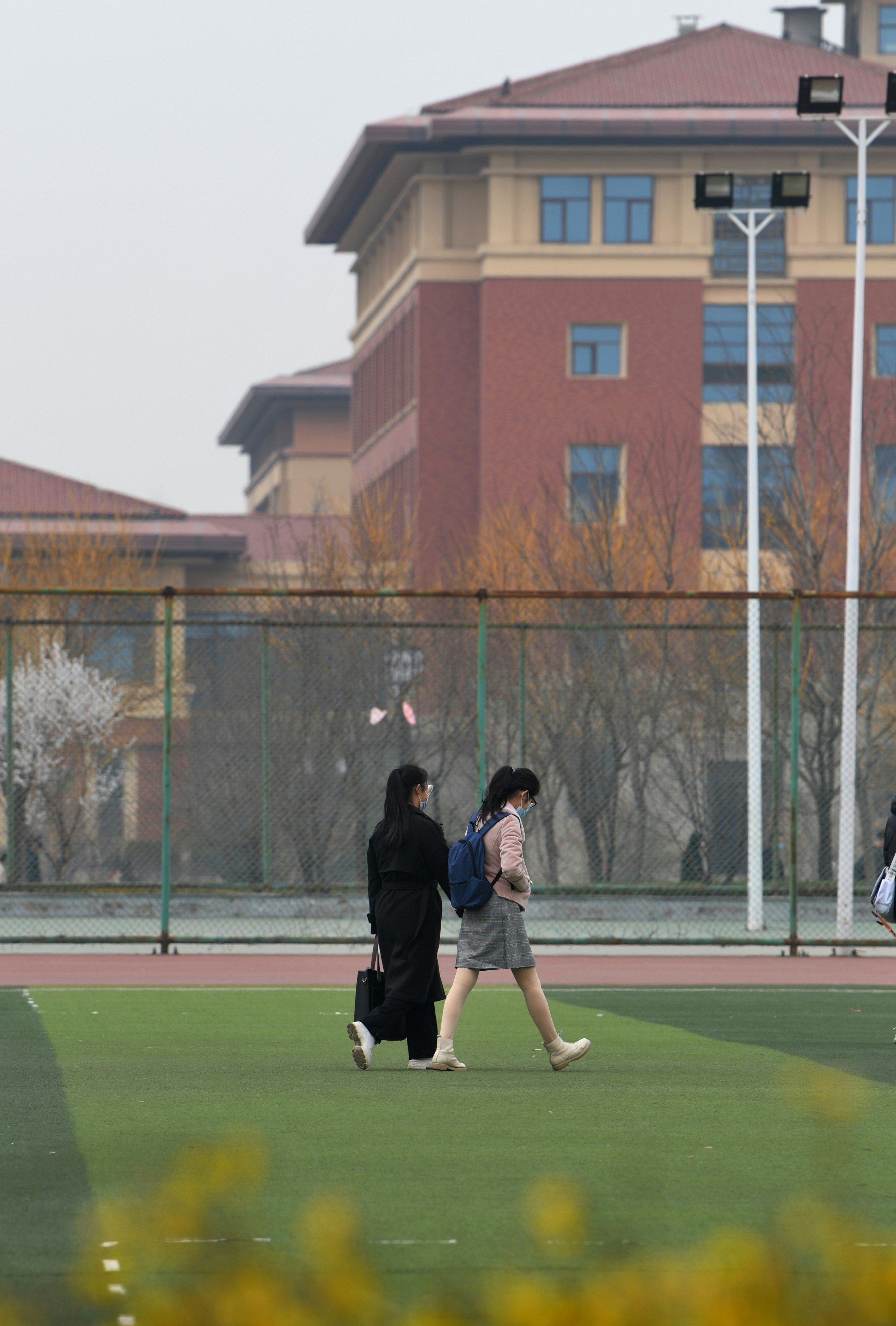 Two Students Crossing the Campus Field with a Brick Building Backdrop
