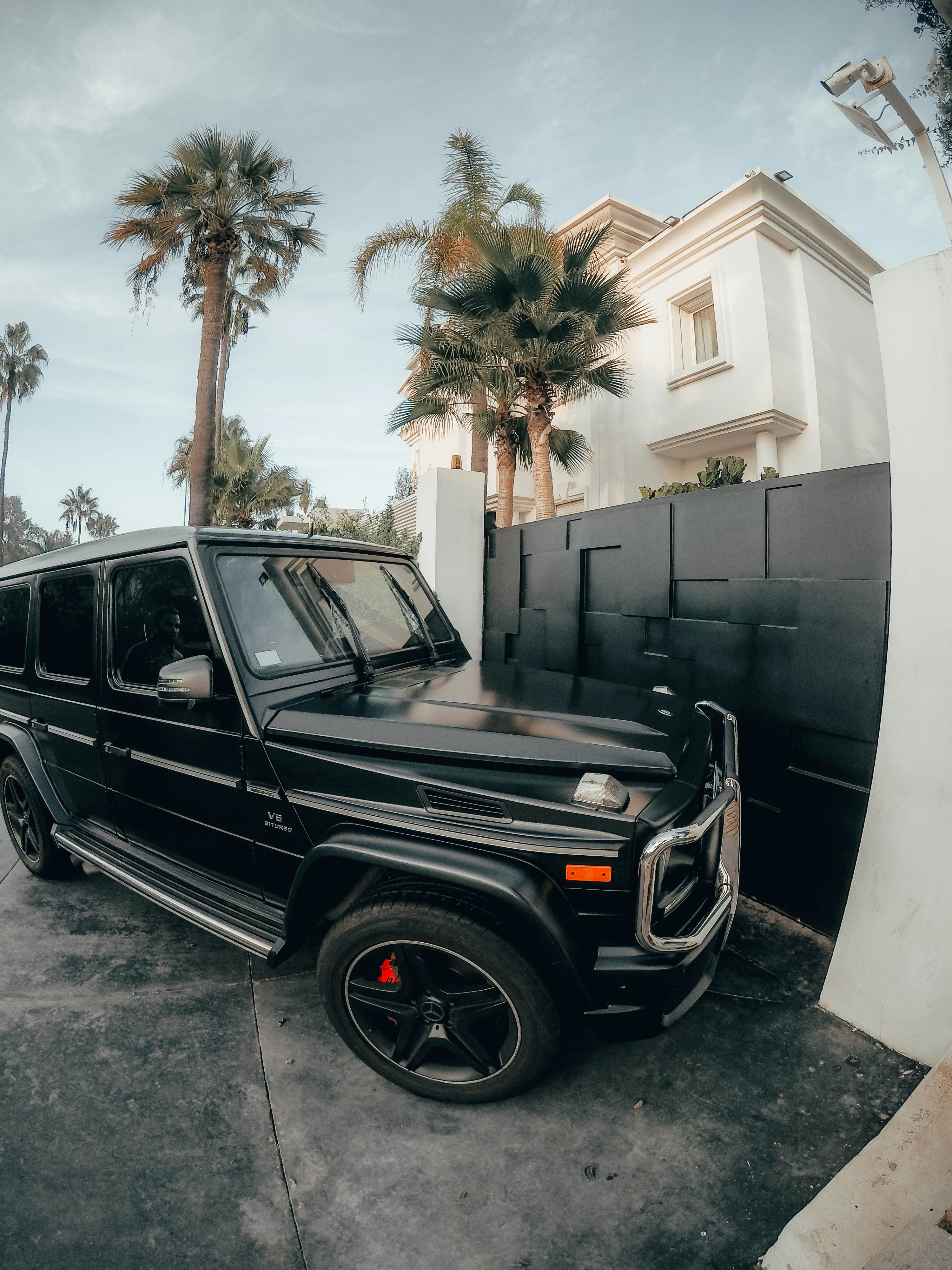 Black Mercedes G-Class parked in a modern driveway surrounded by palm trees and contemporary architecture.