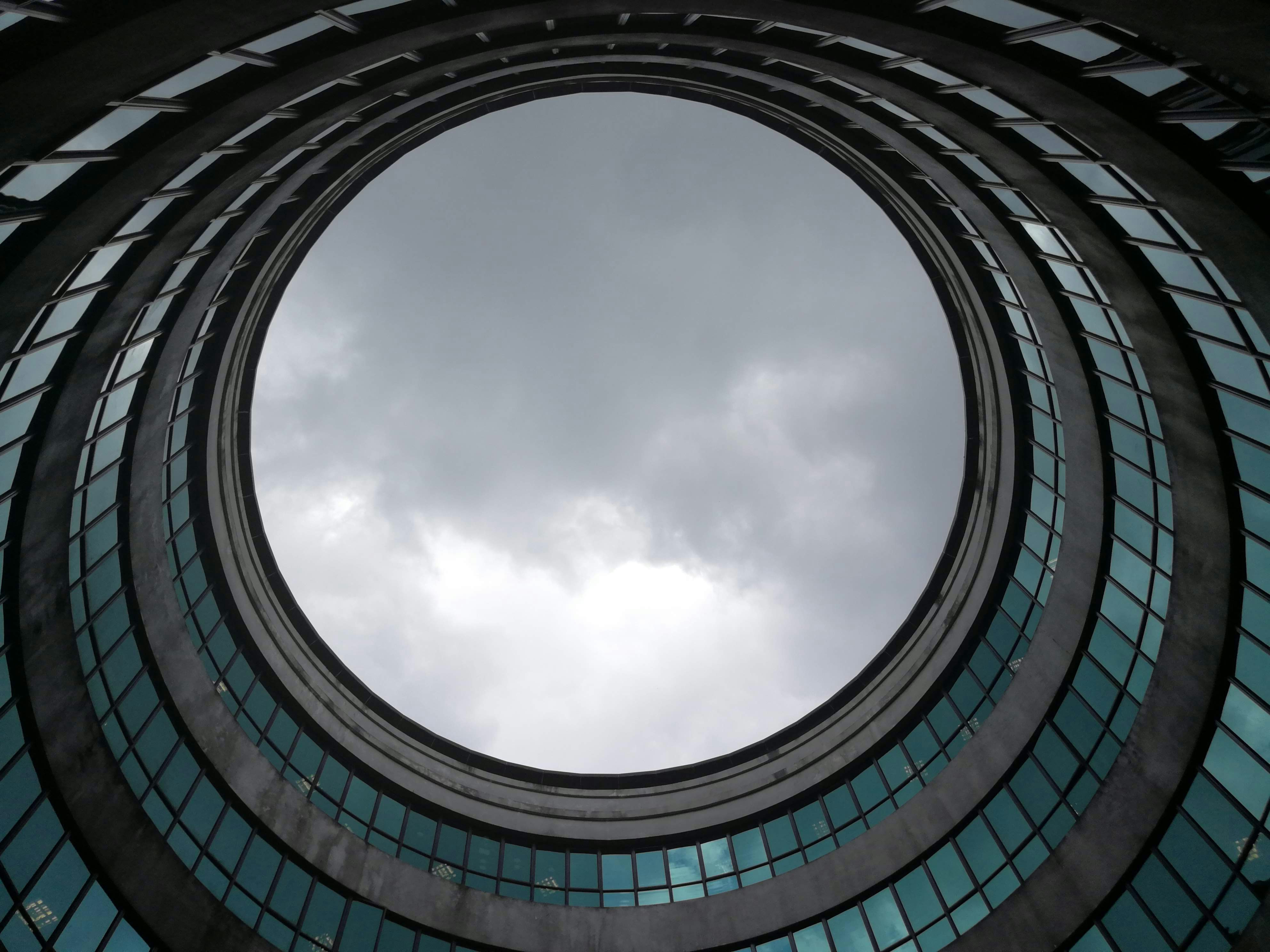 A photograph of a circular glass-and-steel atrium viewed from below, with concentric rings framing a cloudy sky.
