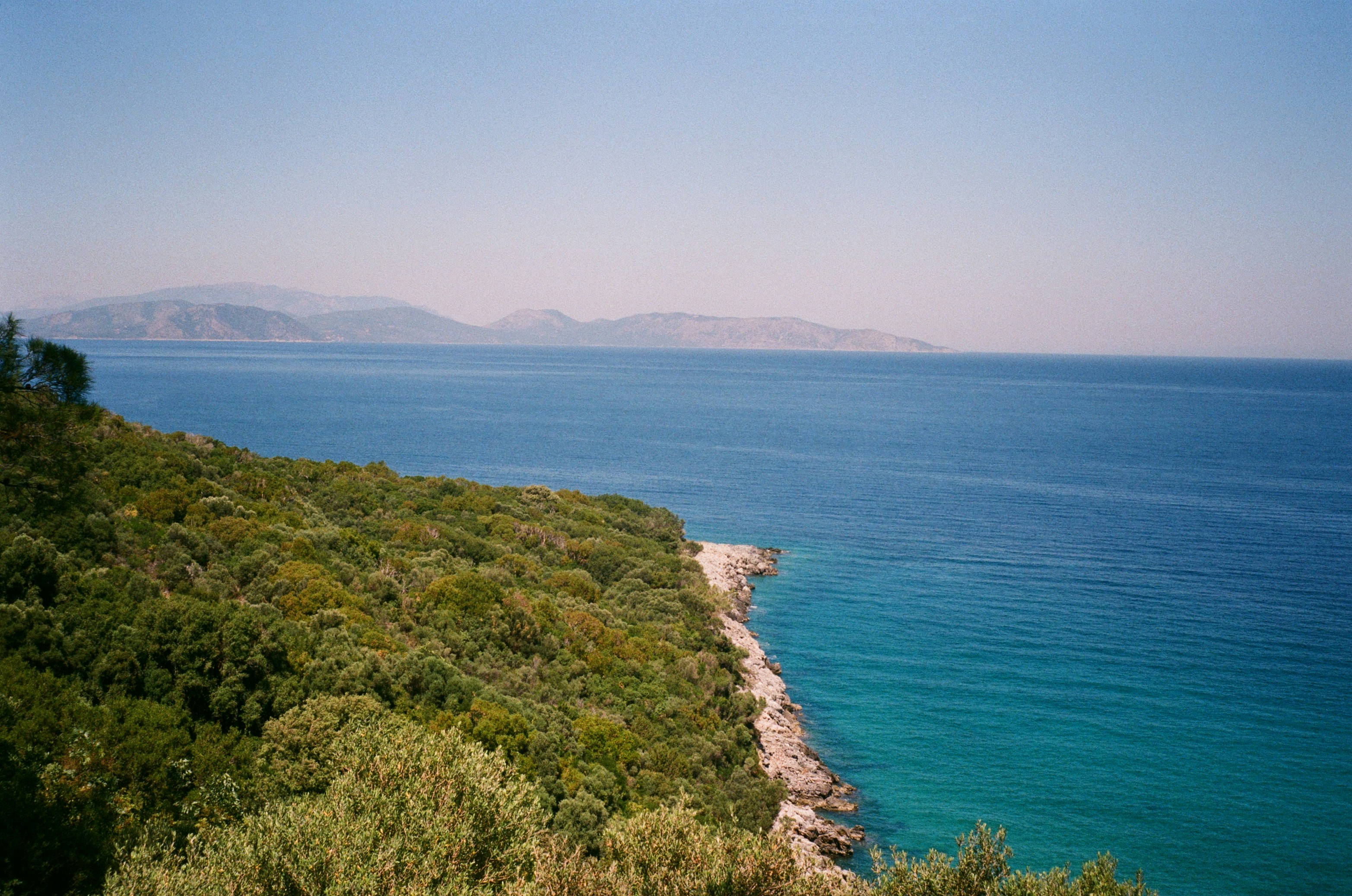 Coastal landscape photograph showing a forested promontory descending to a rocky shoreline where turquoise waters meet the horizon, with distant hills on the far shore.