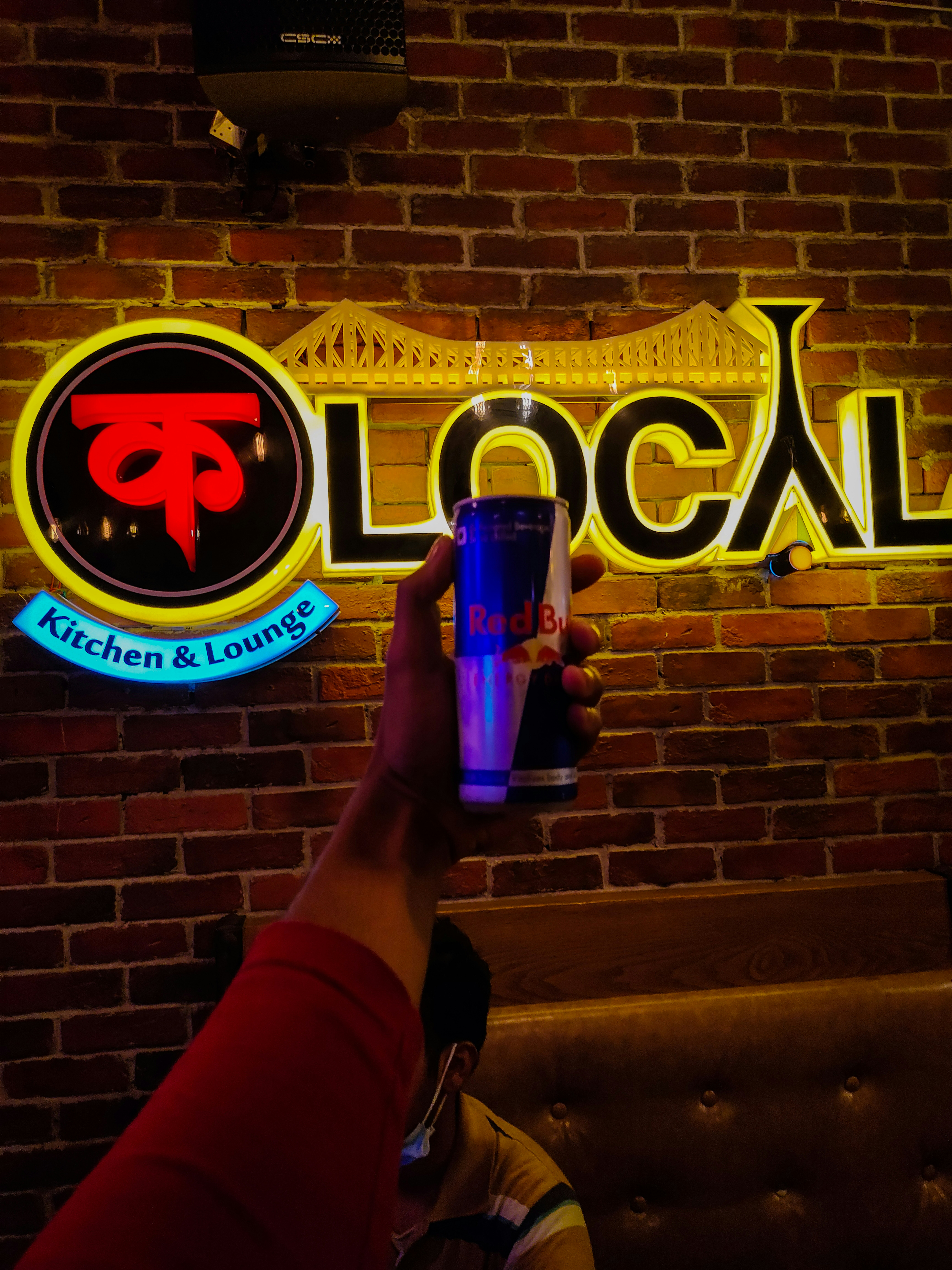 A hand holds a Red Bull can in front of a brick wall illuminated by a bright Local neon sign. The shot emphasizes ambient lighting and bold color contrast in a lively bar-cafe setting.