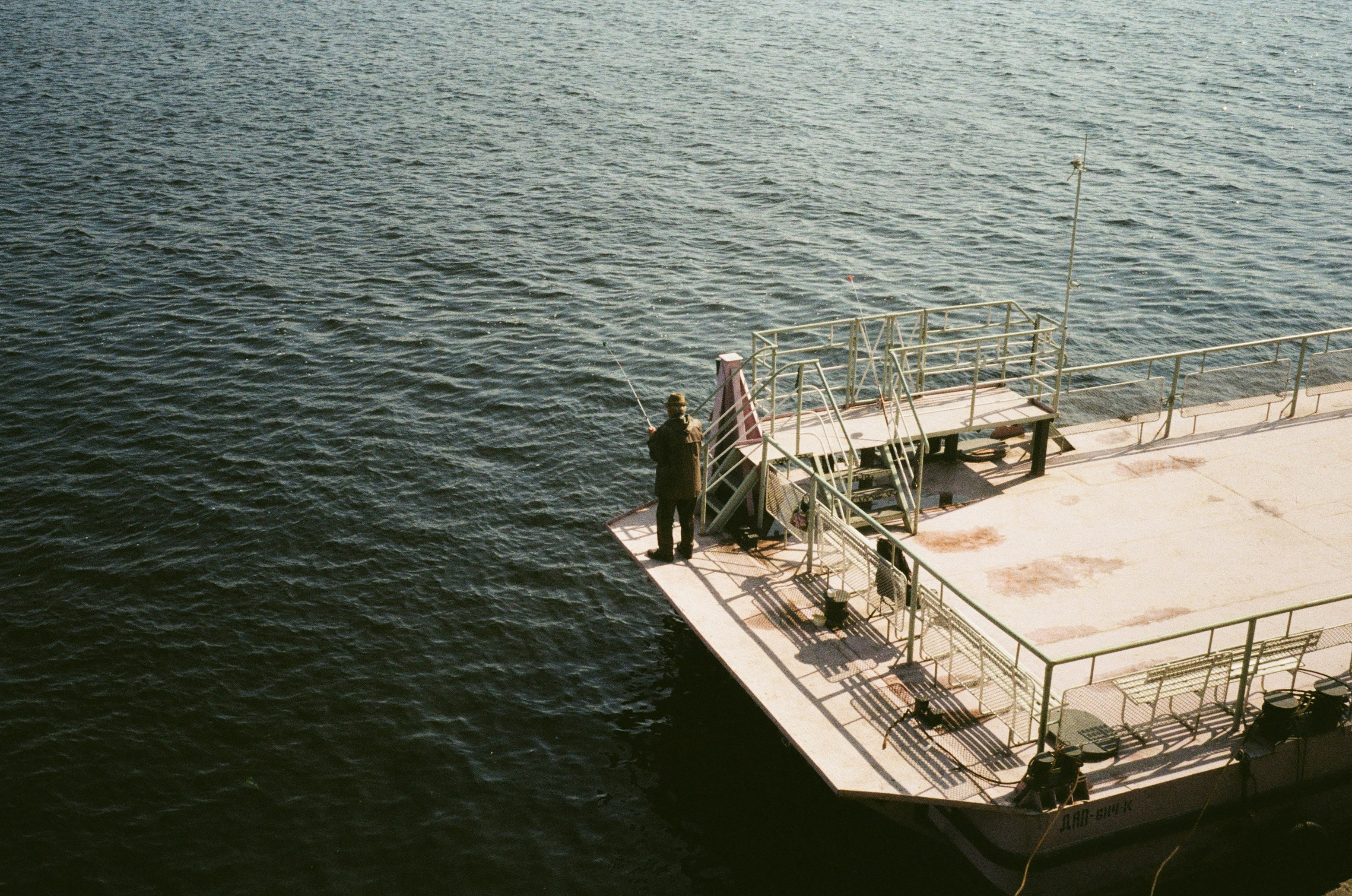 A lone figure stands at the edge of a weathered metal pier extending into calm water, framed by railings and the open sea. The scene emphasizes solitude and the geometric lines of the platform against the expansive backdrop.