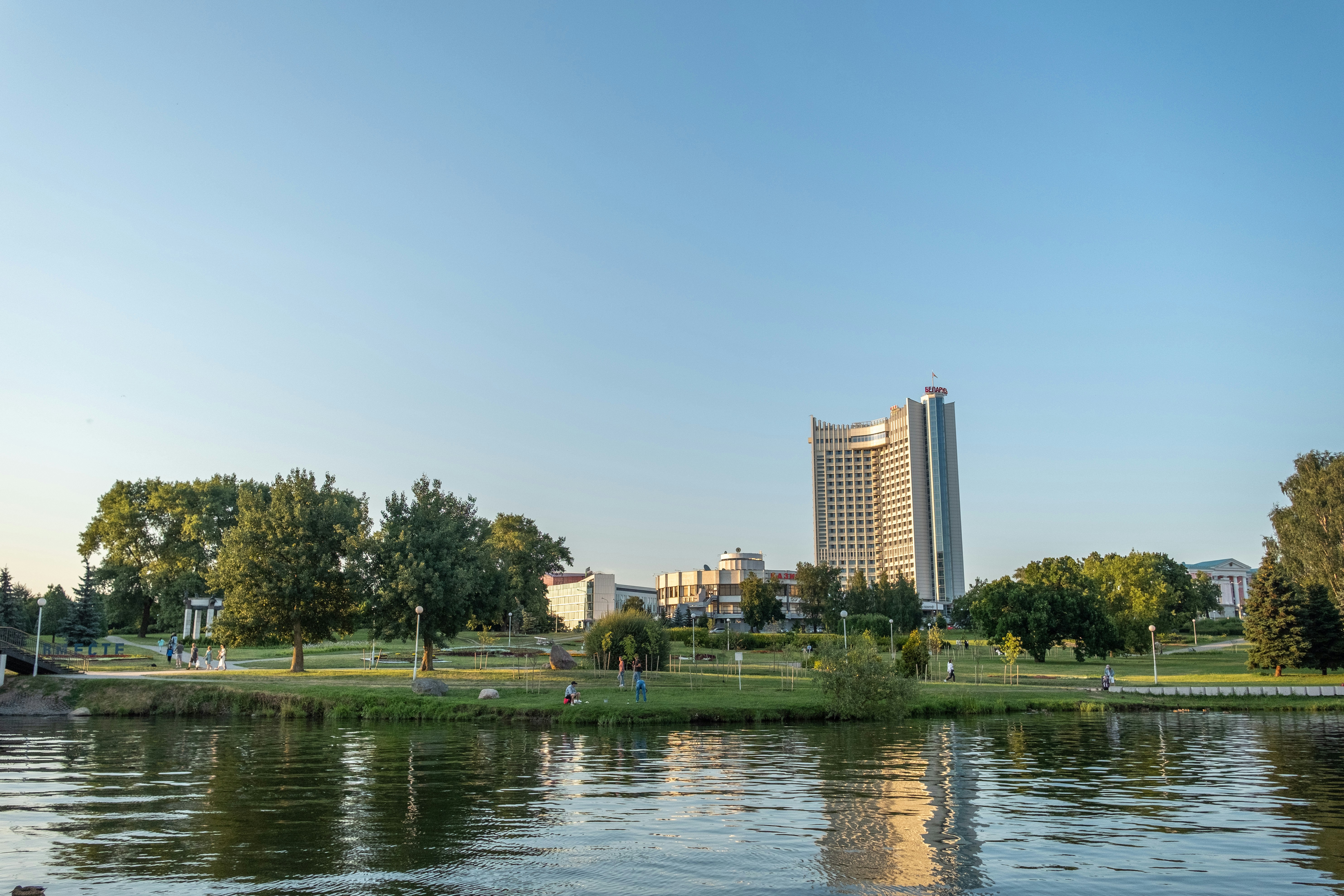 Modern high-rise building alongside a lush park with people enjoying the waterfront. Reflections shimmer on the water's surface.